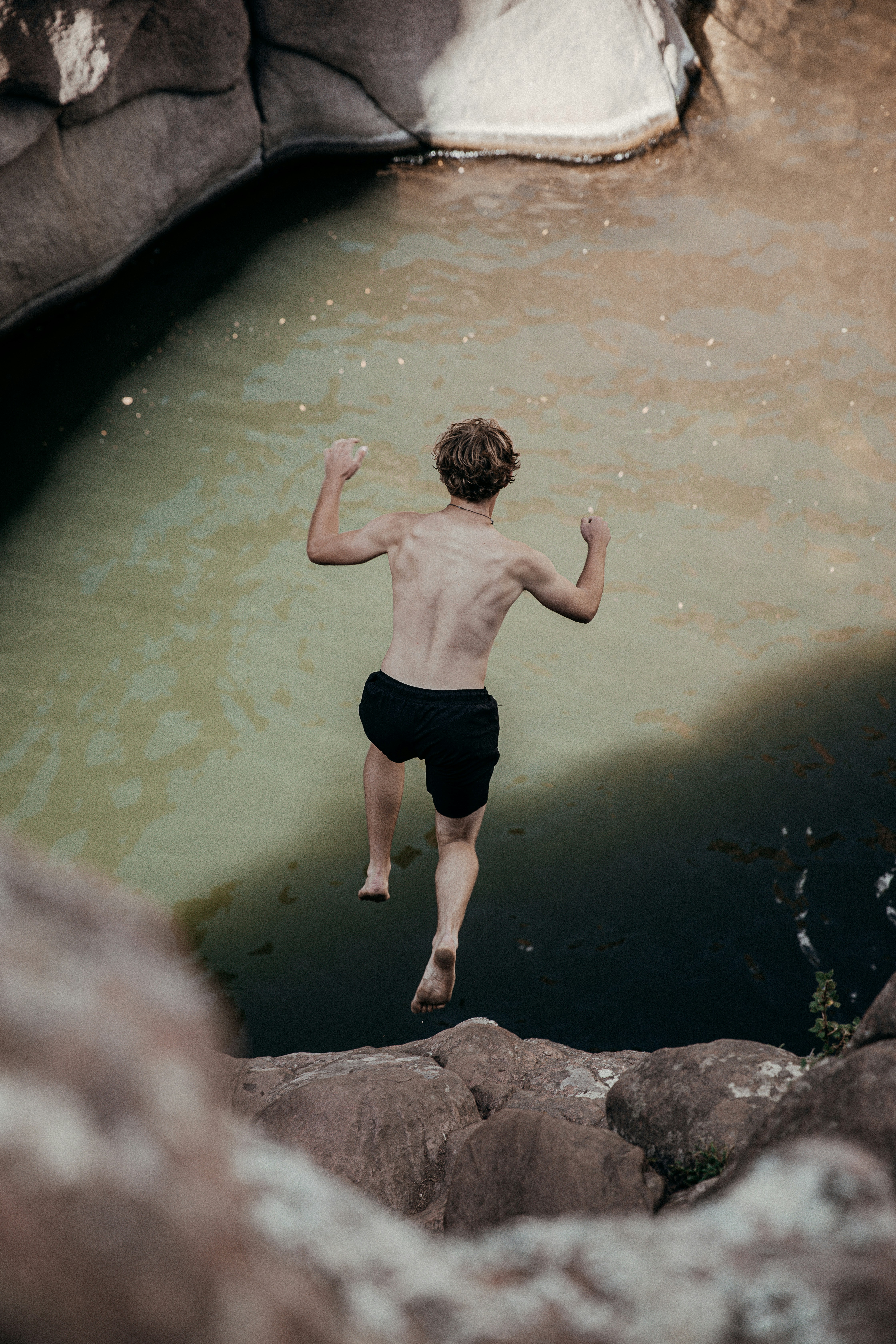 Person in black shorts jumping into a rocky pool from above.