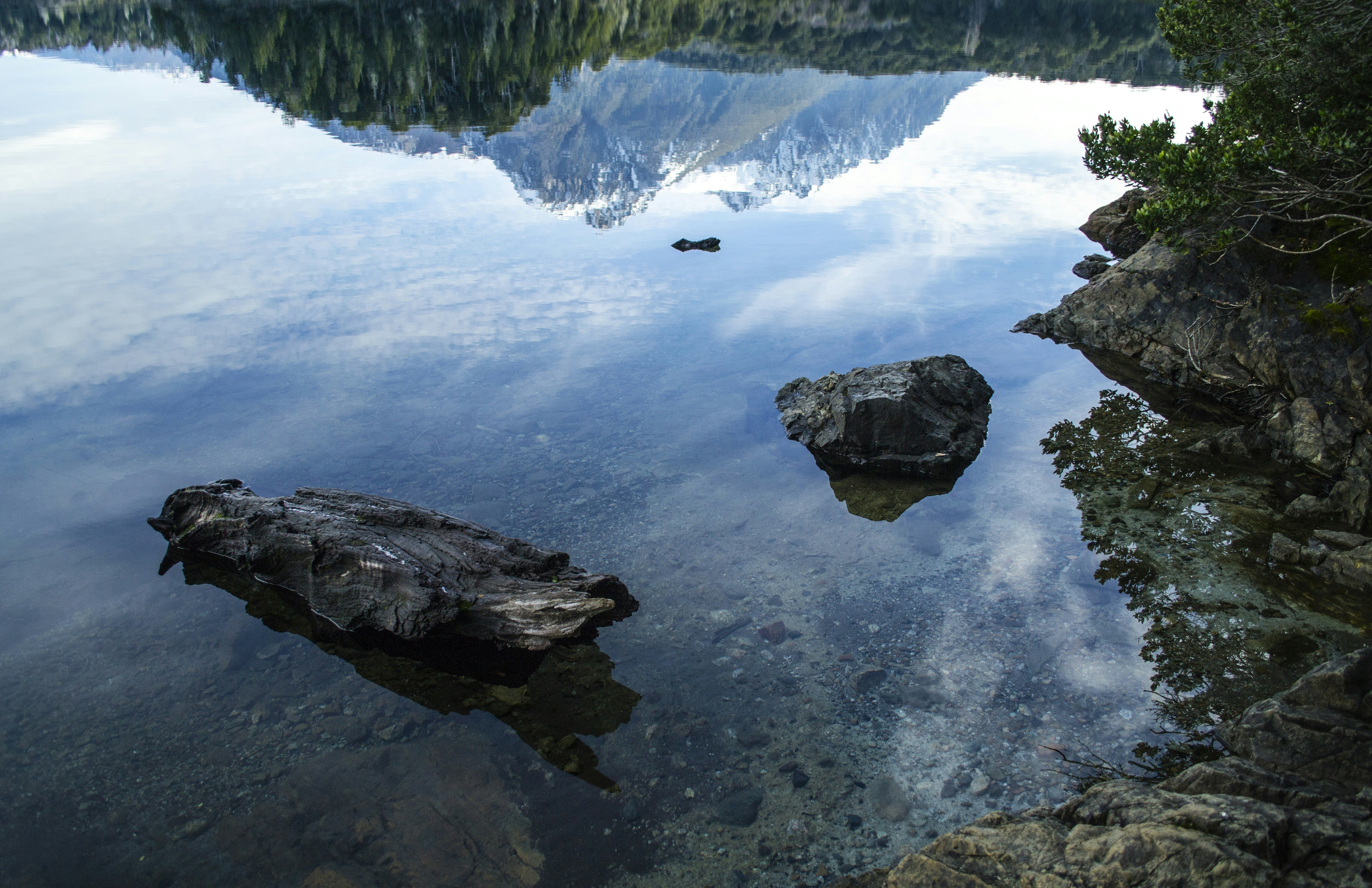 Calm mountain lake reflecting distant peaks and surrounding foliage under a clear sky.
