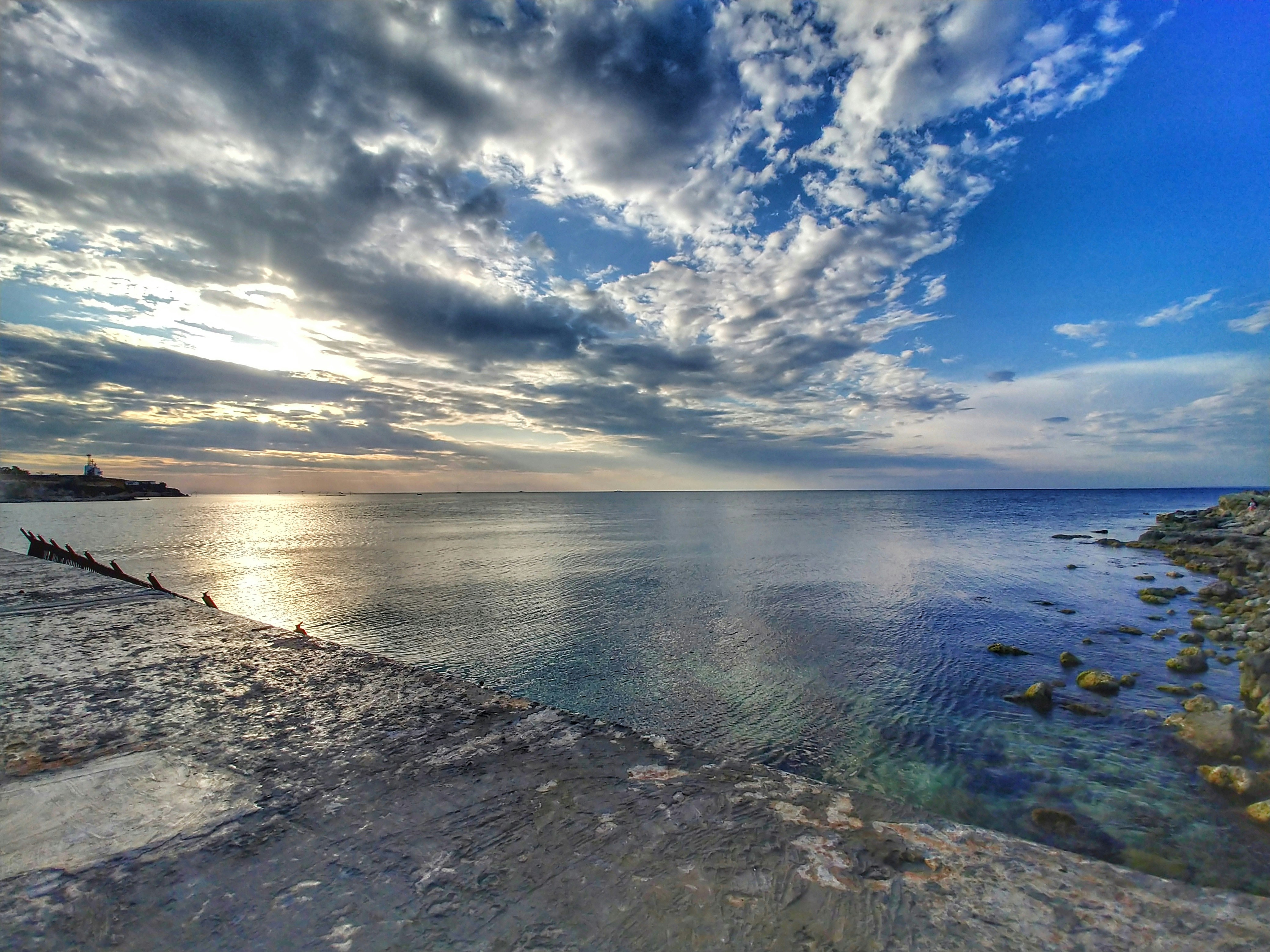 Calm ocean scene with a rocky shoreline under a dynamic sky, reflecting soft hues of blue and gray. The horizon stretches into the distance.