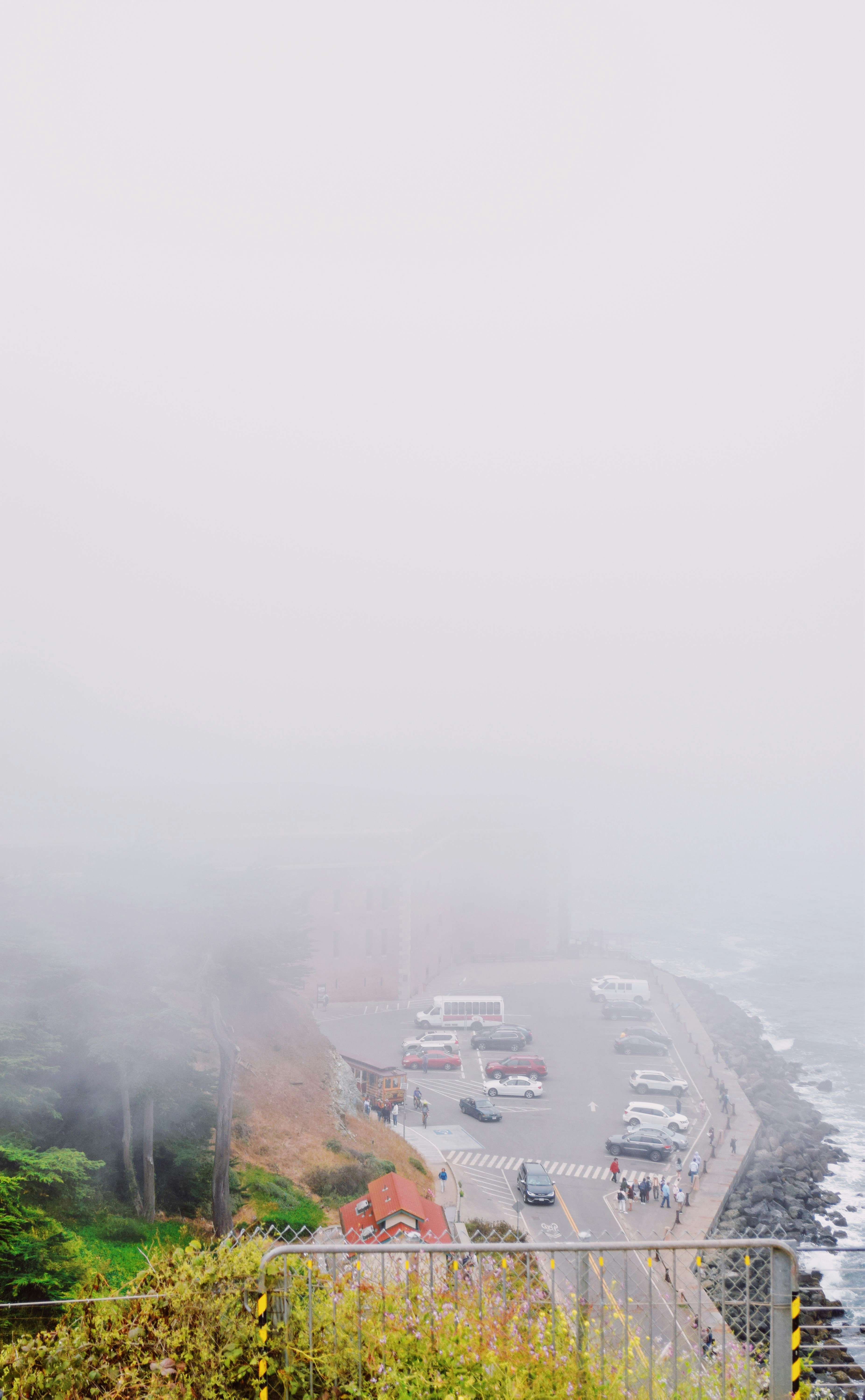 Fog enveloping a coastal parking area, with vehicles and people visible along the shore. The scene captures the tranquil yet mysterious atmosphere of a foggy day.