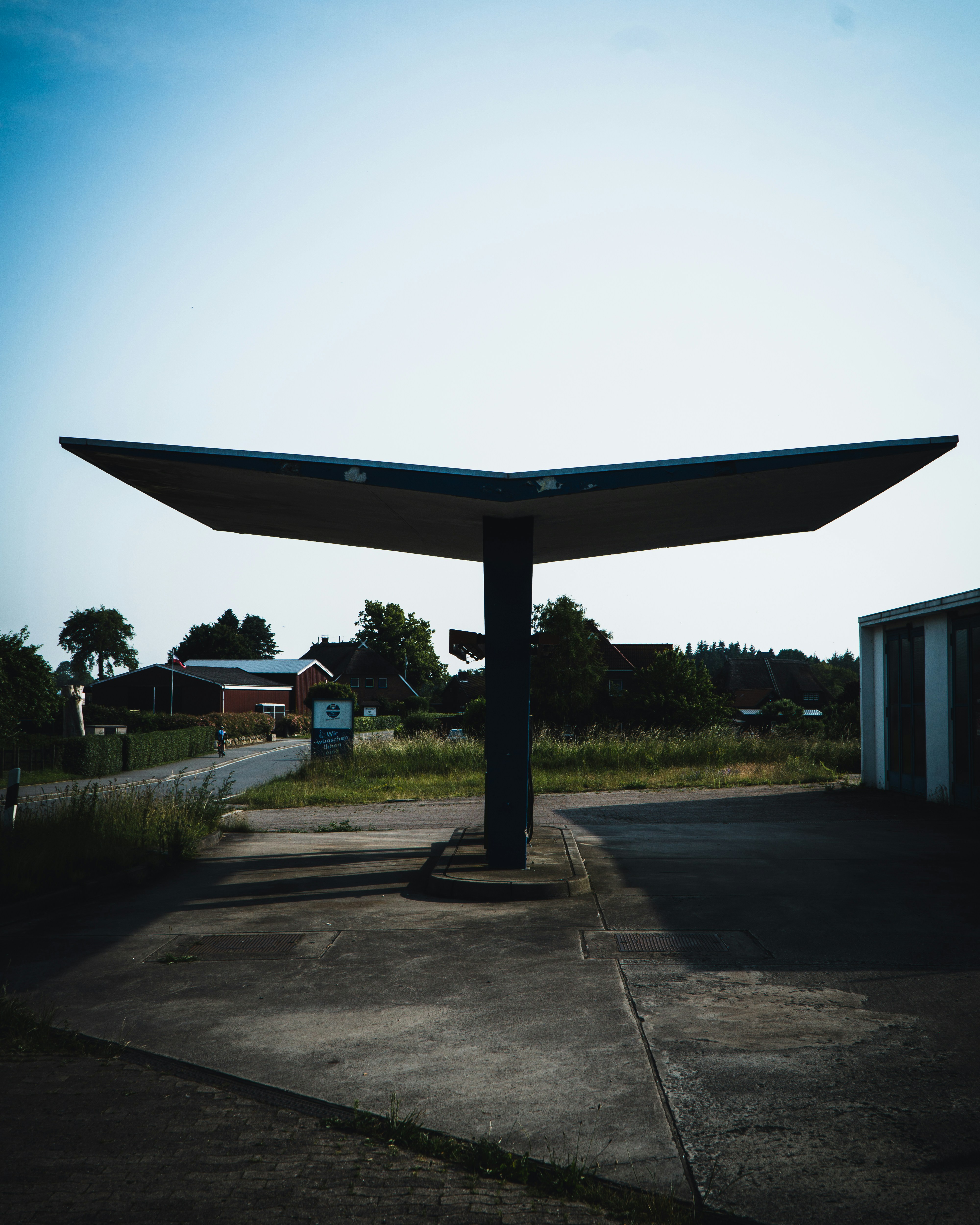An isolated gas station canopy stands against a clear sky, surrounded by overgrown grass and distant buildings, evoking a sense of neglect.