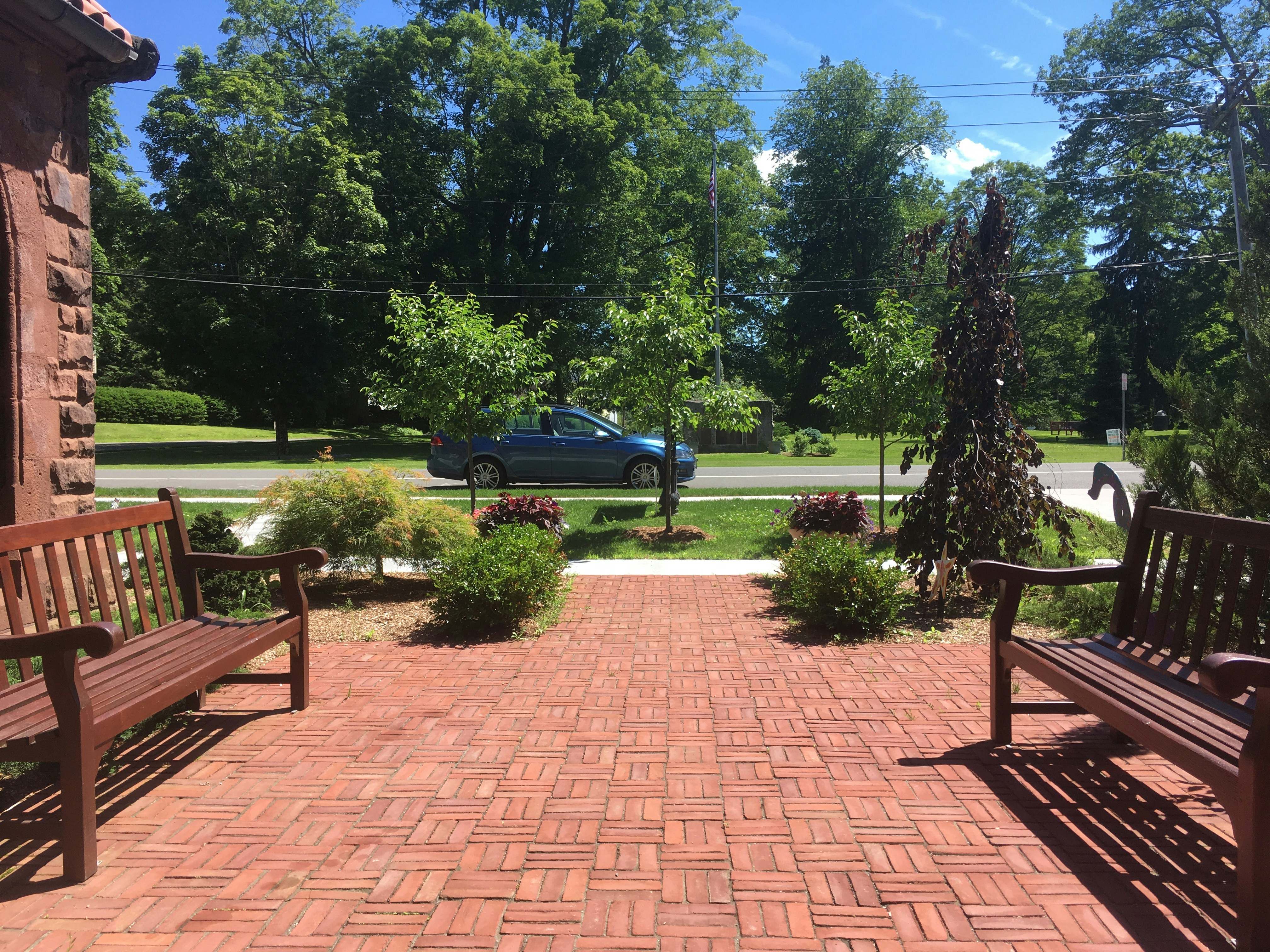 Two wooden benches face each other on a sunlit brick patio, framed by lush greenery and a passing blue car.