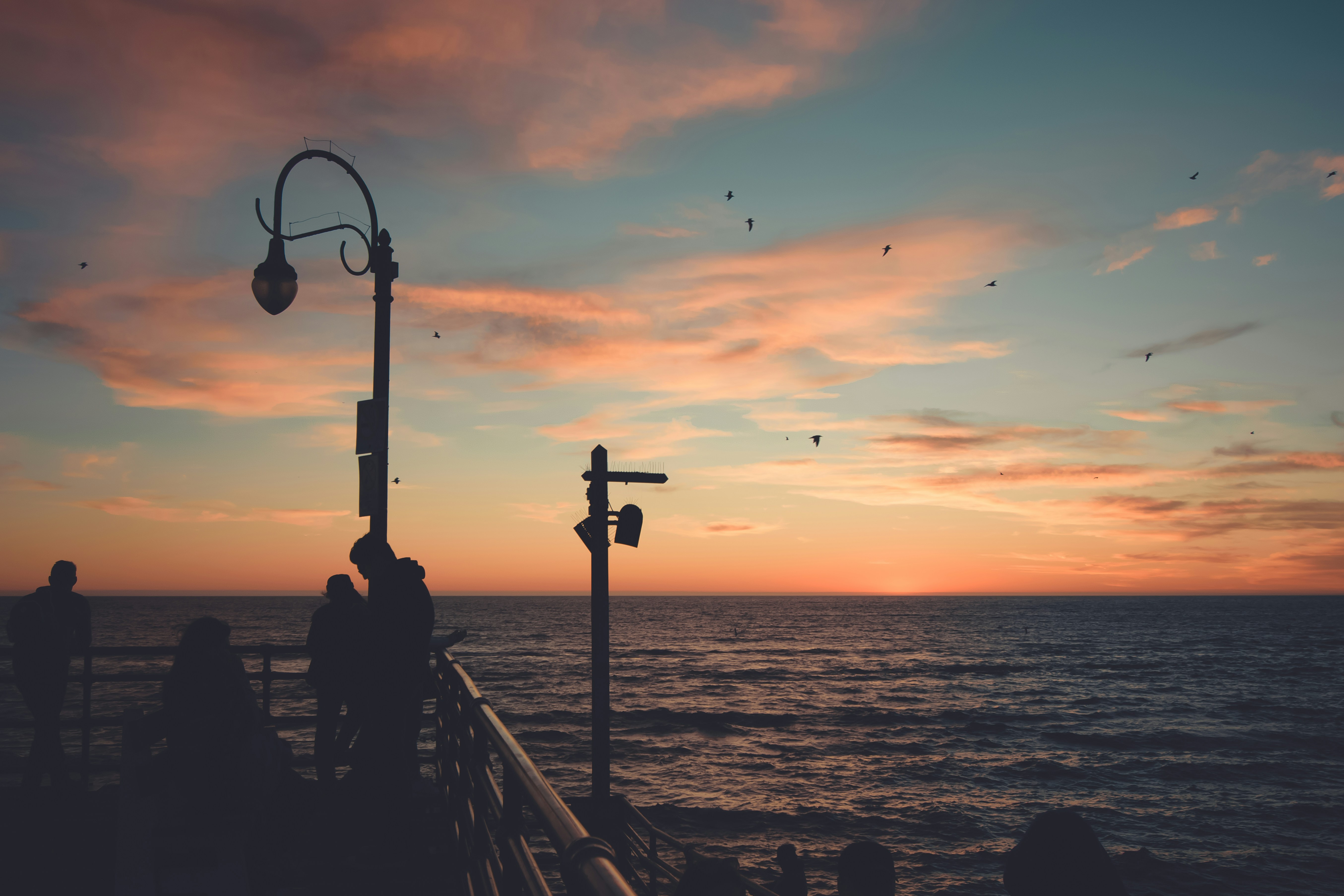 Silhouetted figures converse on a pier as the sun sets over the ocean, with a colorful sky scattered with clouds and birds in flight.