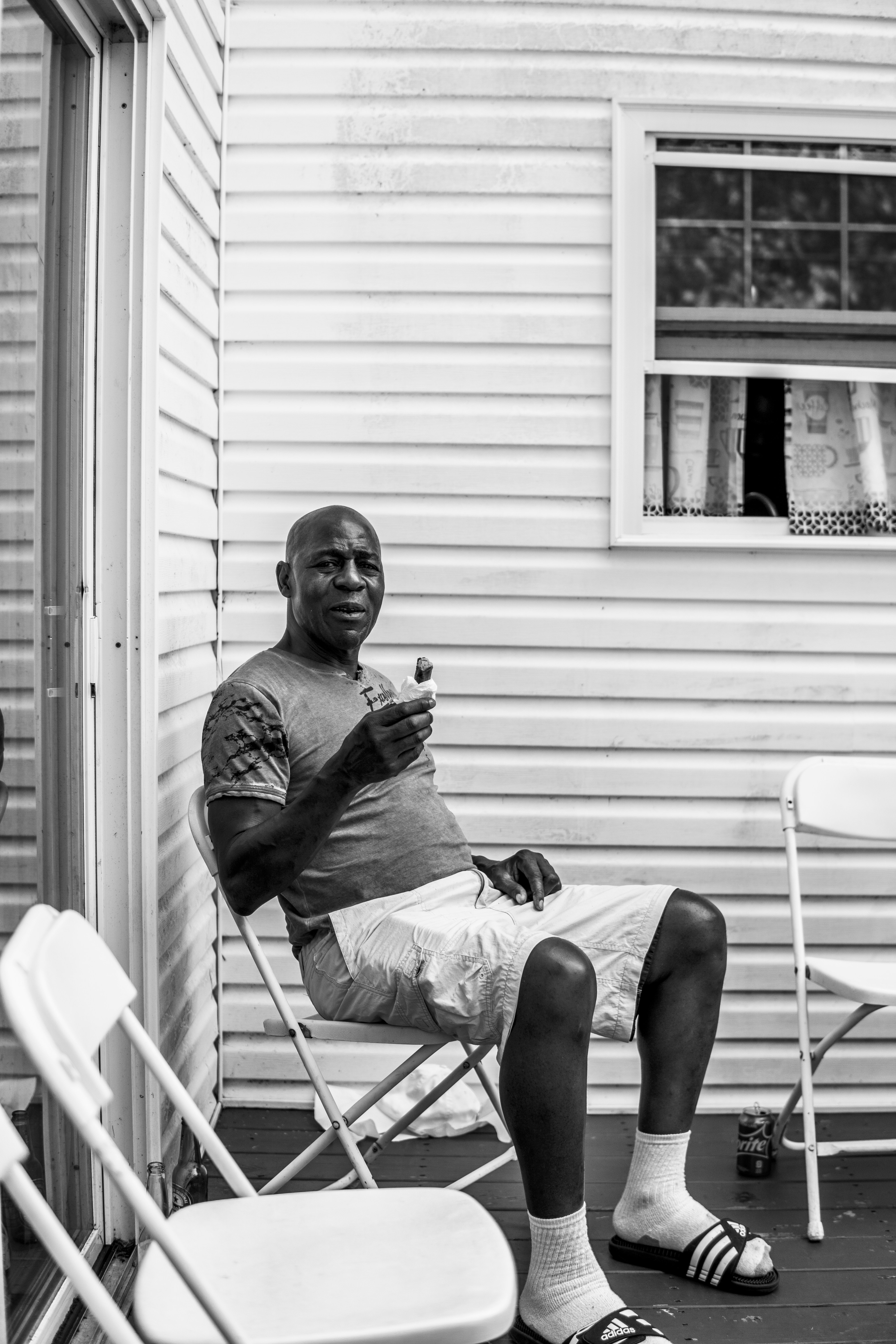 Man seated on a chair, enjoying a snack on a porch with a white wooden backdrop. The scene captures a relaxed afternoon atmosphere.