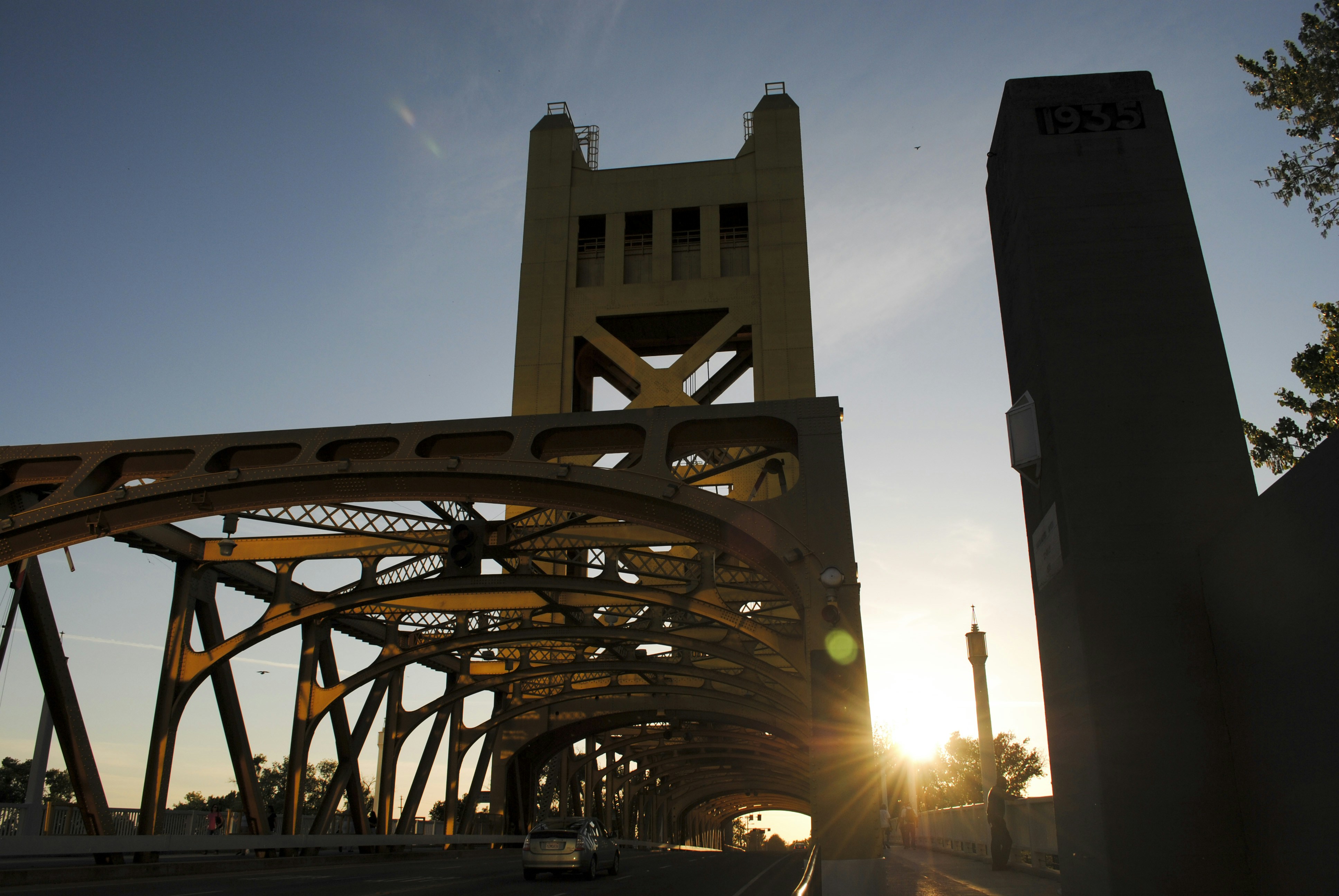 Silhouette of a metal bridge tower against a sunset sky, with the sun peeking through the structure.