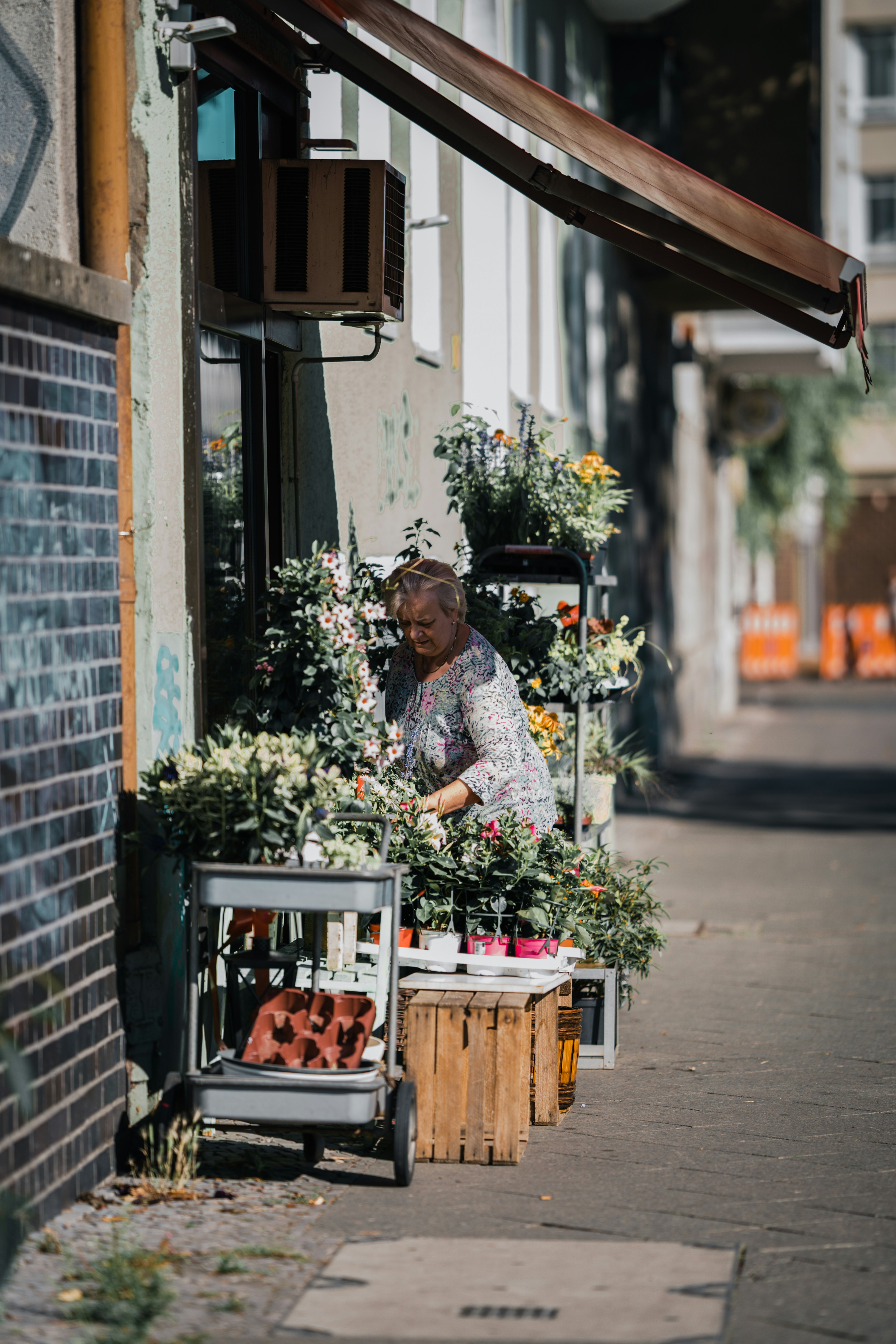 A florist arranges vibrant flowers outside her shop, surrounded by greenery and rustic crates. The scene captures the essence of a bustling urban market.