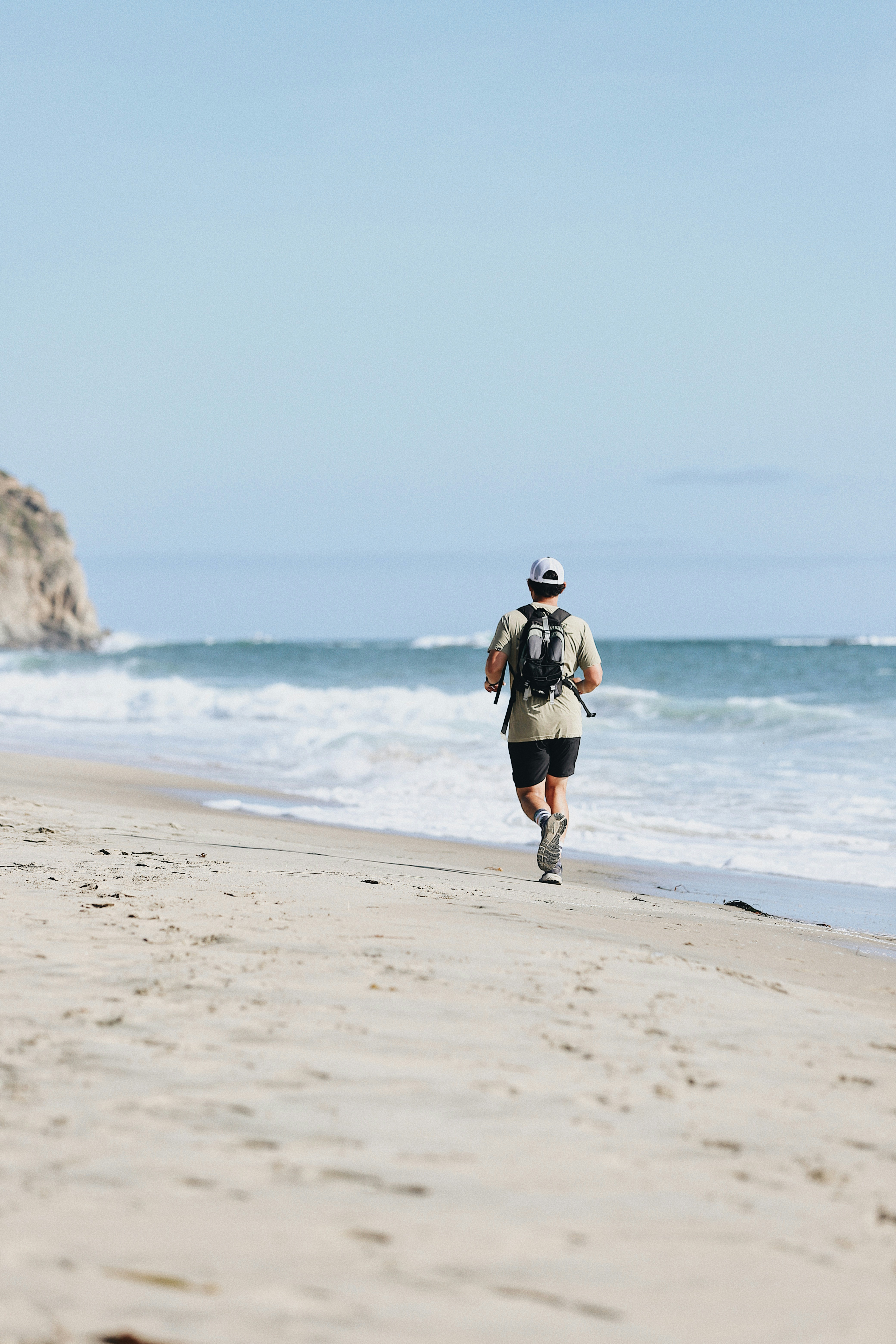 A lone runner strides along a sandy beach, with gentle waves lapping at the shore under a clear sky.