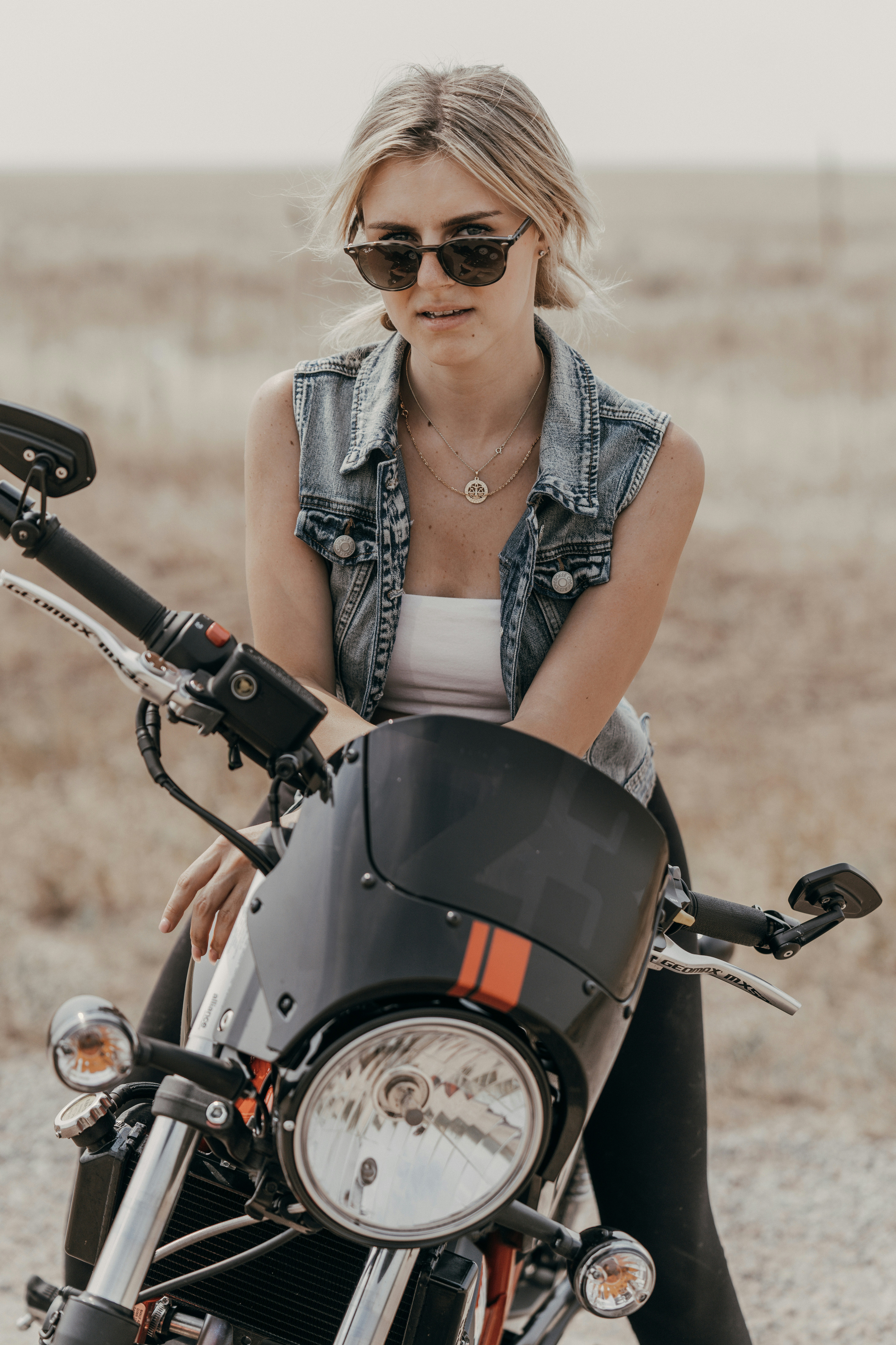 Young woman in a denim vest sits confidently on a motorcycle, exuding a sense of adventure and freedom. The backdrop features a vast, open landscape.
