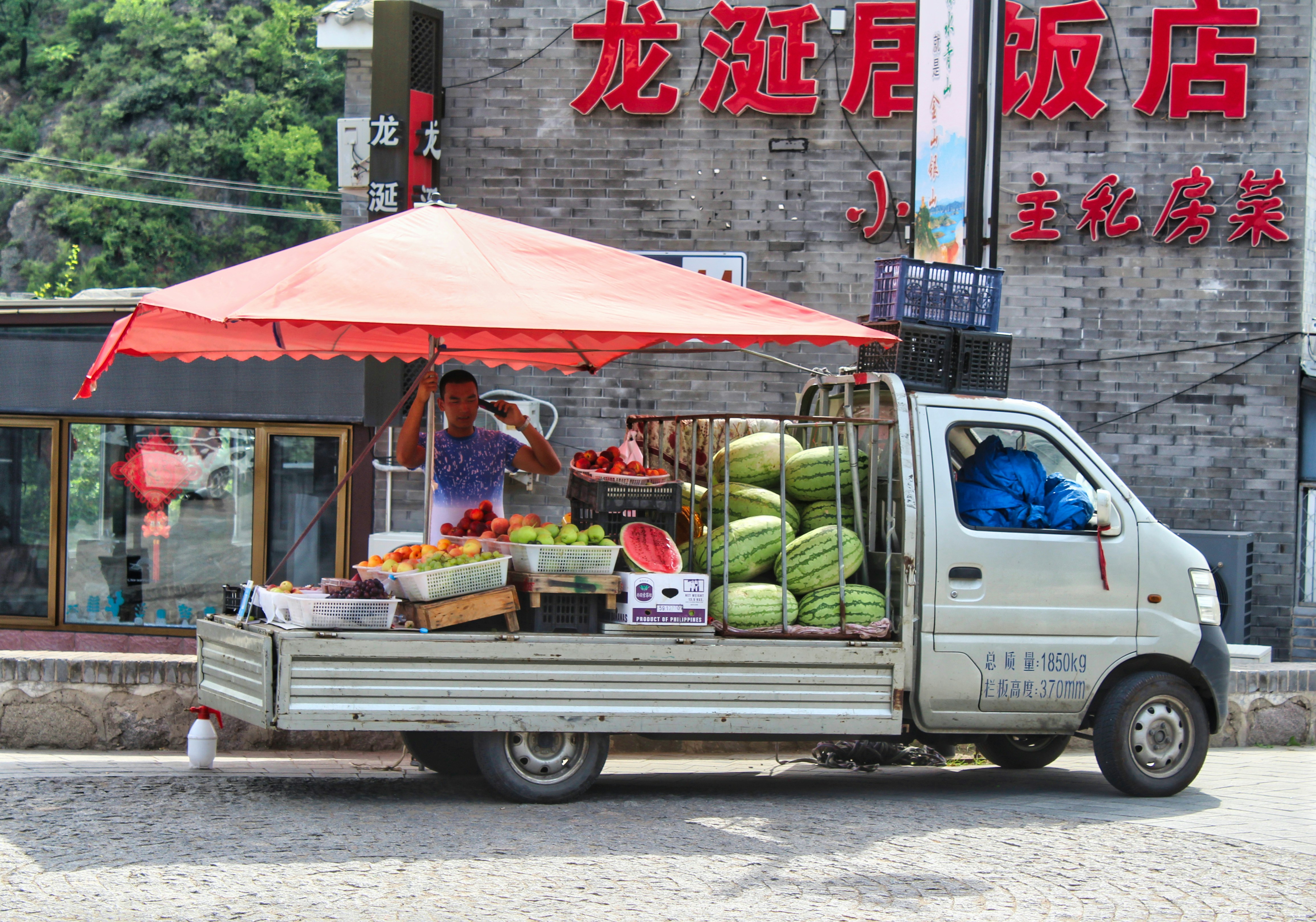 A mobile fruit stand displaying an array of fresh produce, including watermelons and apples, under a red canopy. A vendor engages with customers in a bustling market scene.
