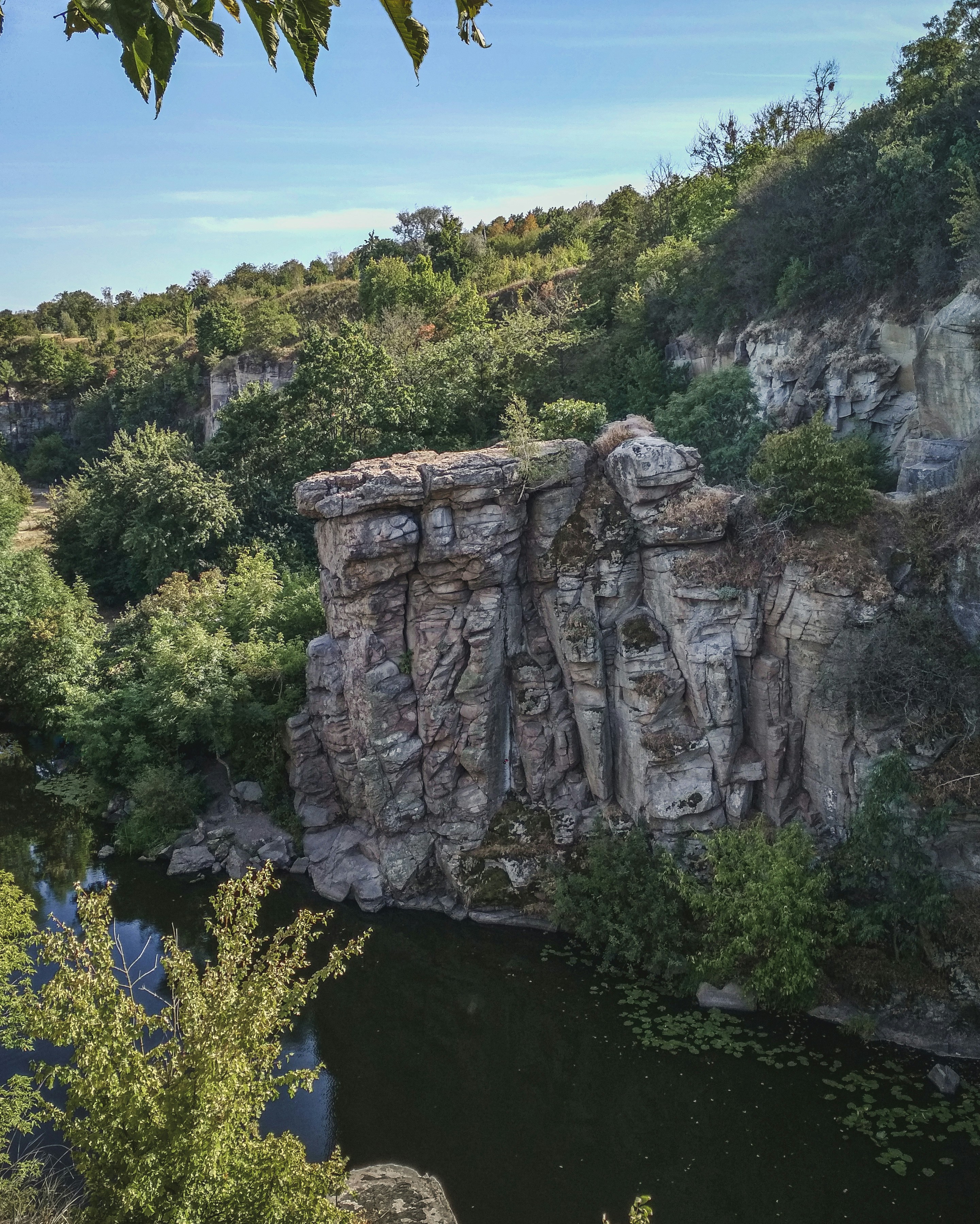 Sunlit limestone pillars rise from a rocky gorge beside a calm river, framed by lush trees under a clear sky.