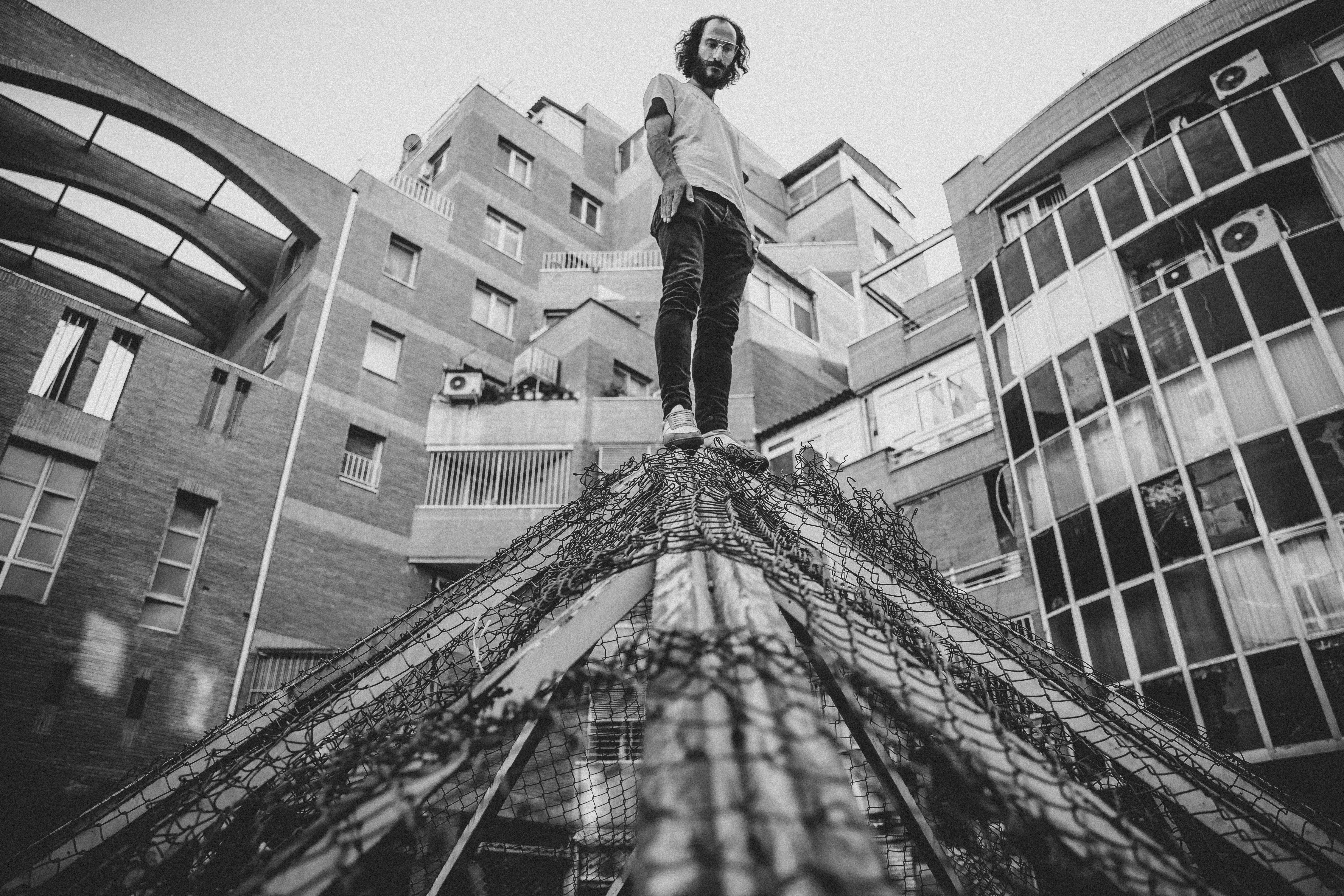 Individual poised atop a metal structure in an urban setting, surrounded by contemporary architecture. The monochrome tones emphasize the stark contrasts of the environment.