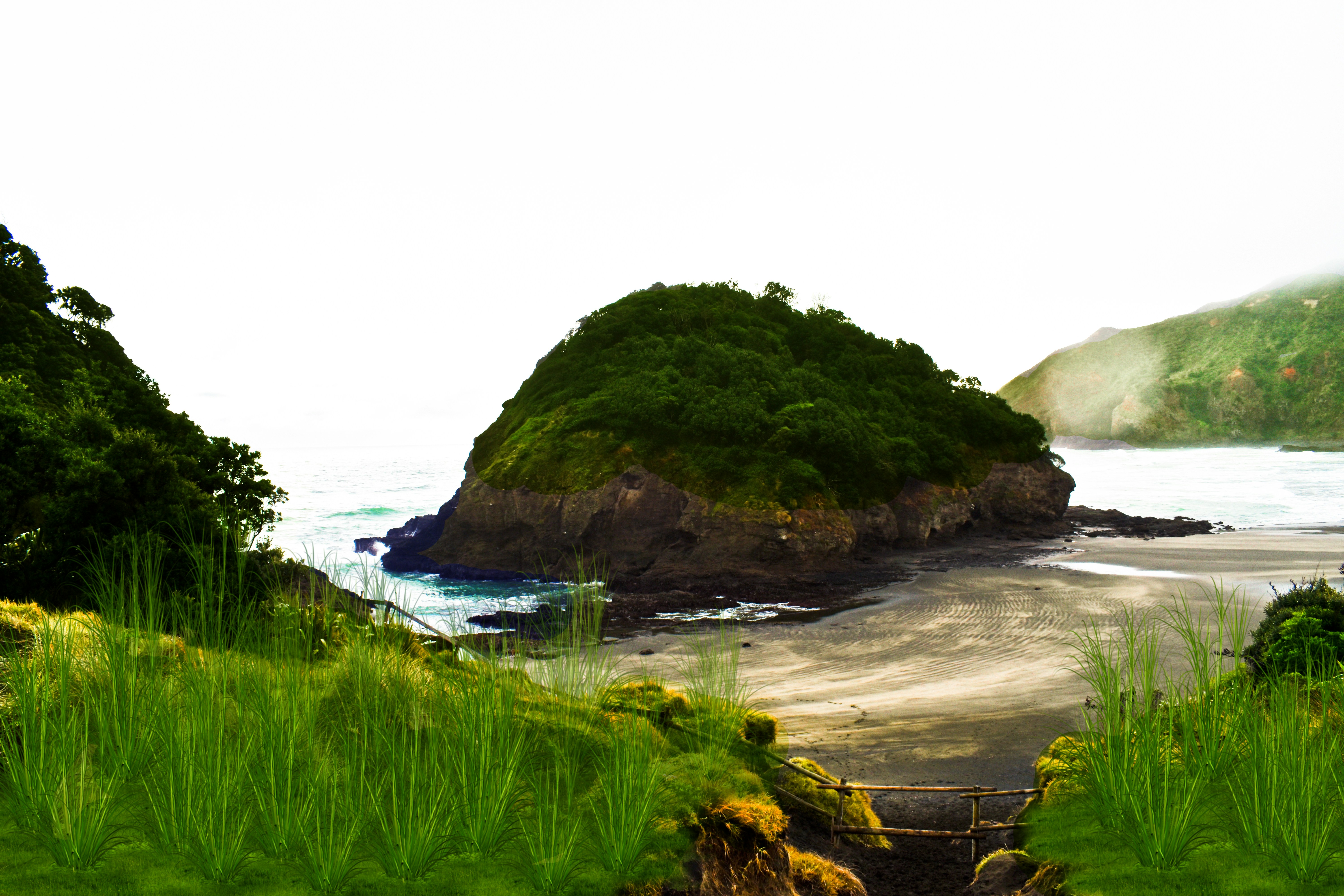 Lush green hills and rocky outcrops frame a secluded beach with gentle waves under a hazy sky.