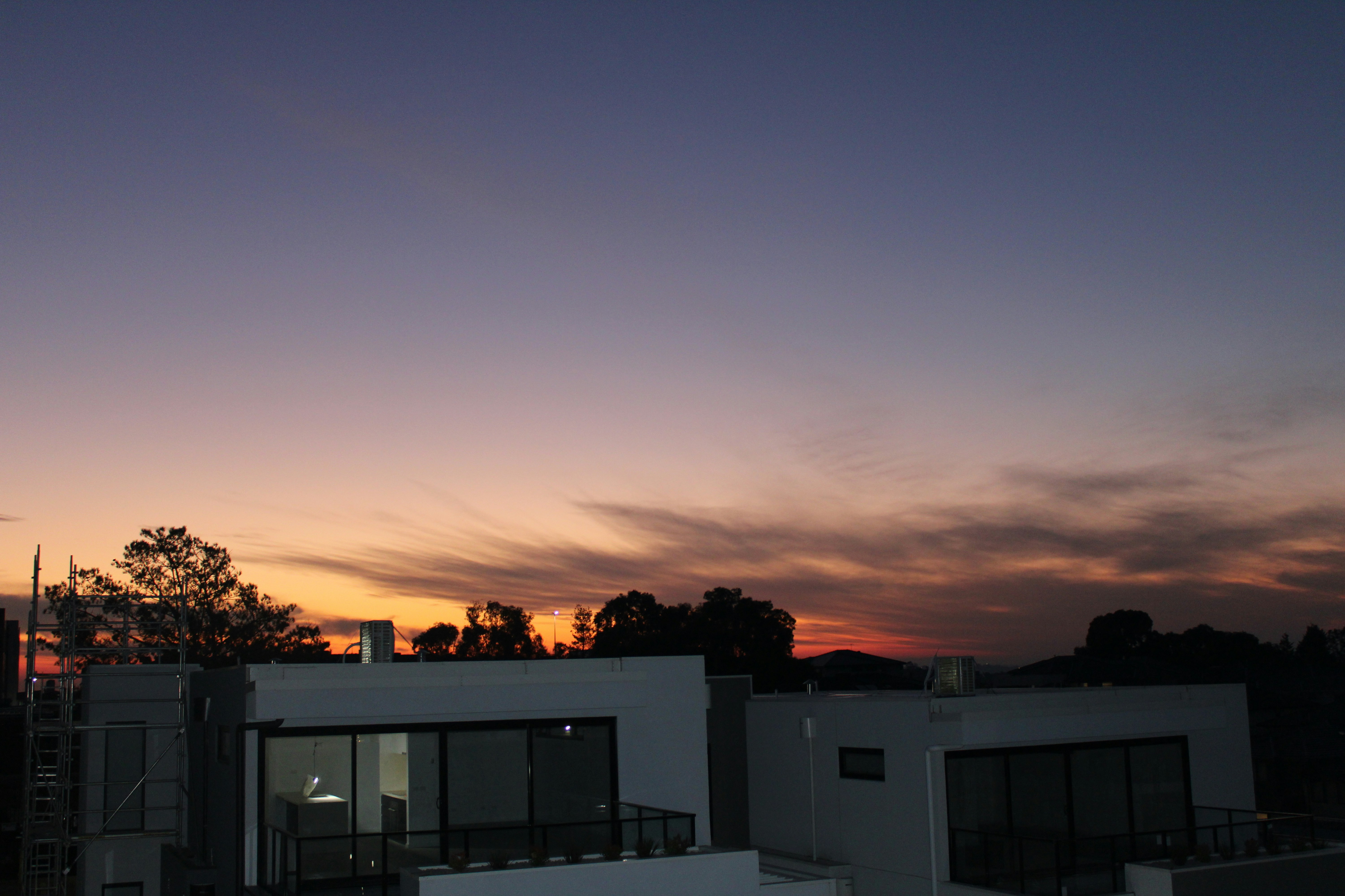 Evening photograph of modern rooftops silhouetted against a warm sunset gradient, with a bright interior window drawing the eye.