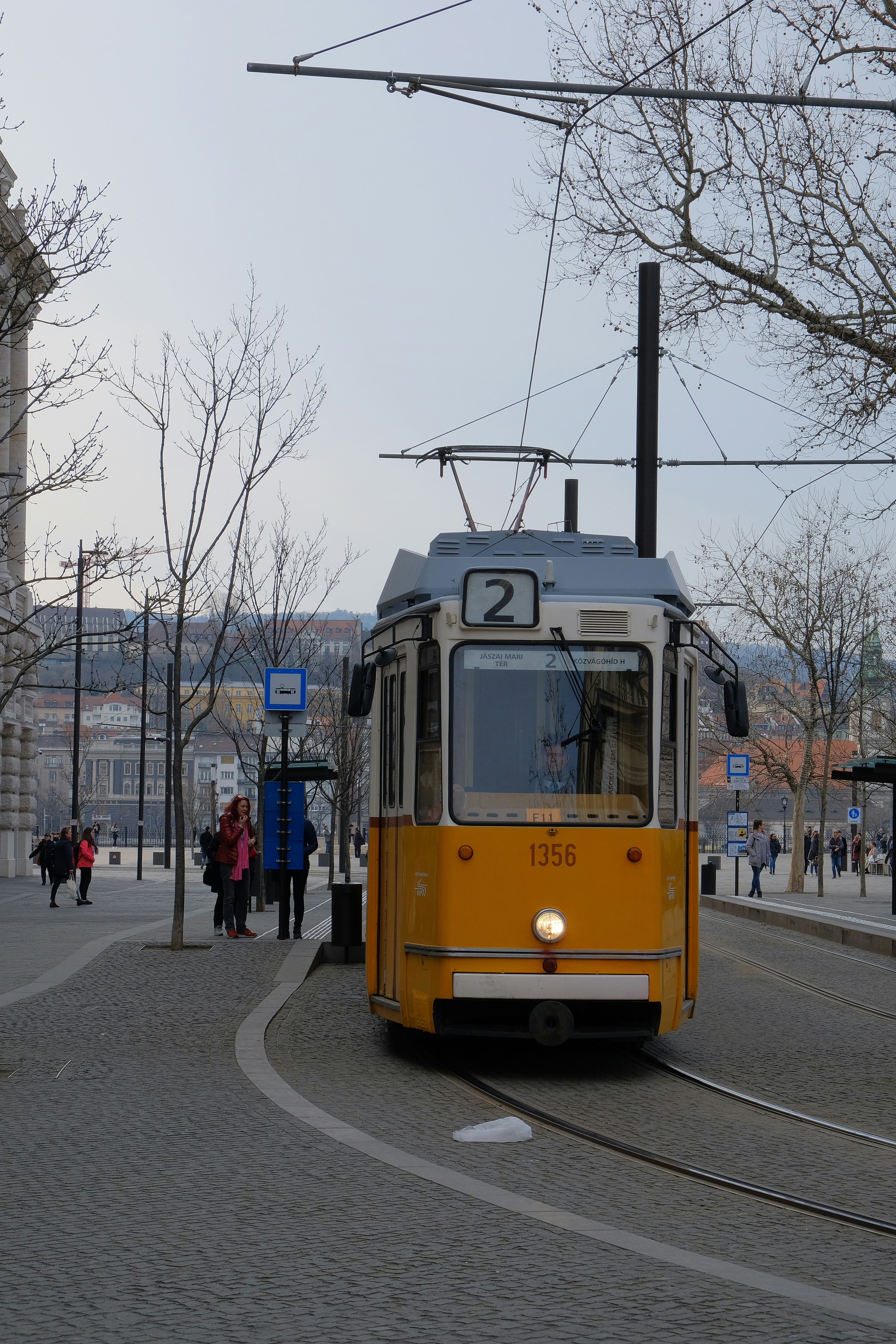 A vintage yellow tram approaches a station amidst a modern urban landscape, with pedestrians waiting nearby.