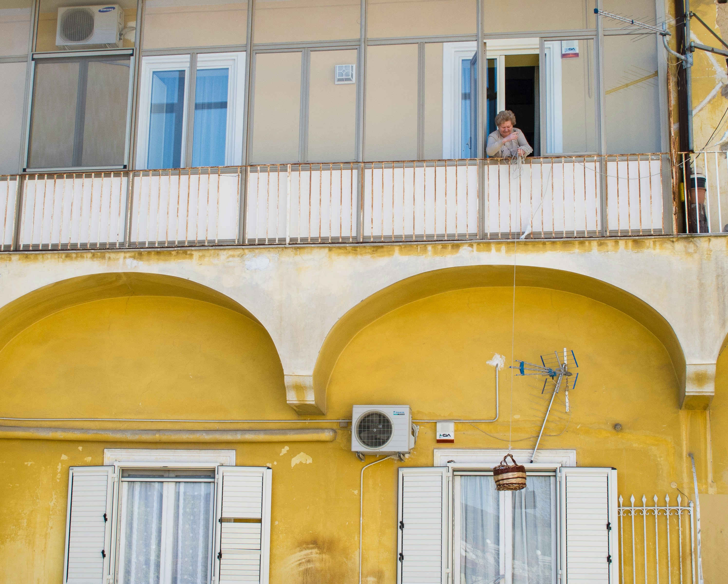 Man gazing from a balcony of a yellow building with arched windows and shutters.