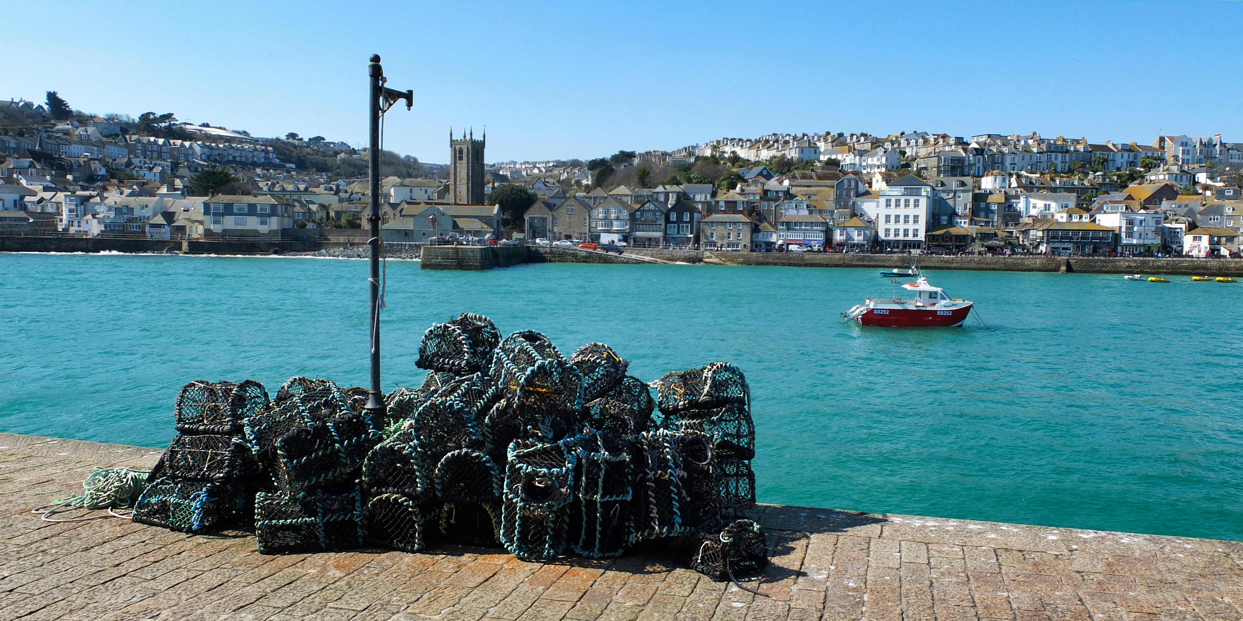 Fishing pots stacked on the quay with a vibrant blue harbor in the background, showcasing a small boat and coastal town.