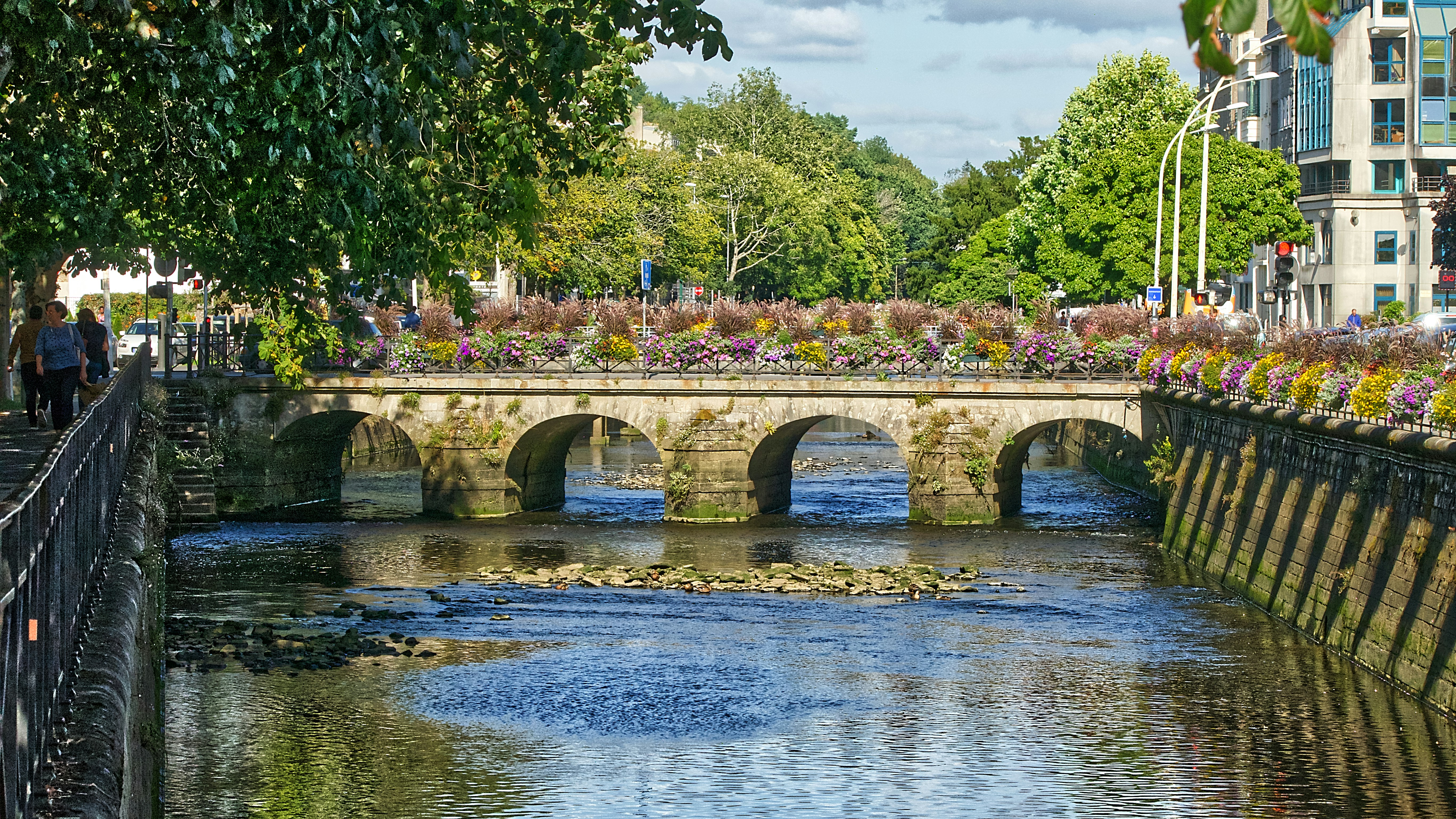 Historic stone bridge adorned with vibrant flowers spans a calm river, surrounded by lush greenery and urban architecture.
