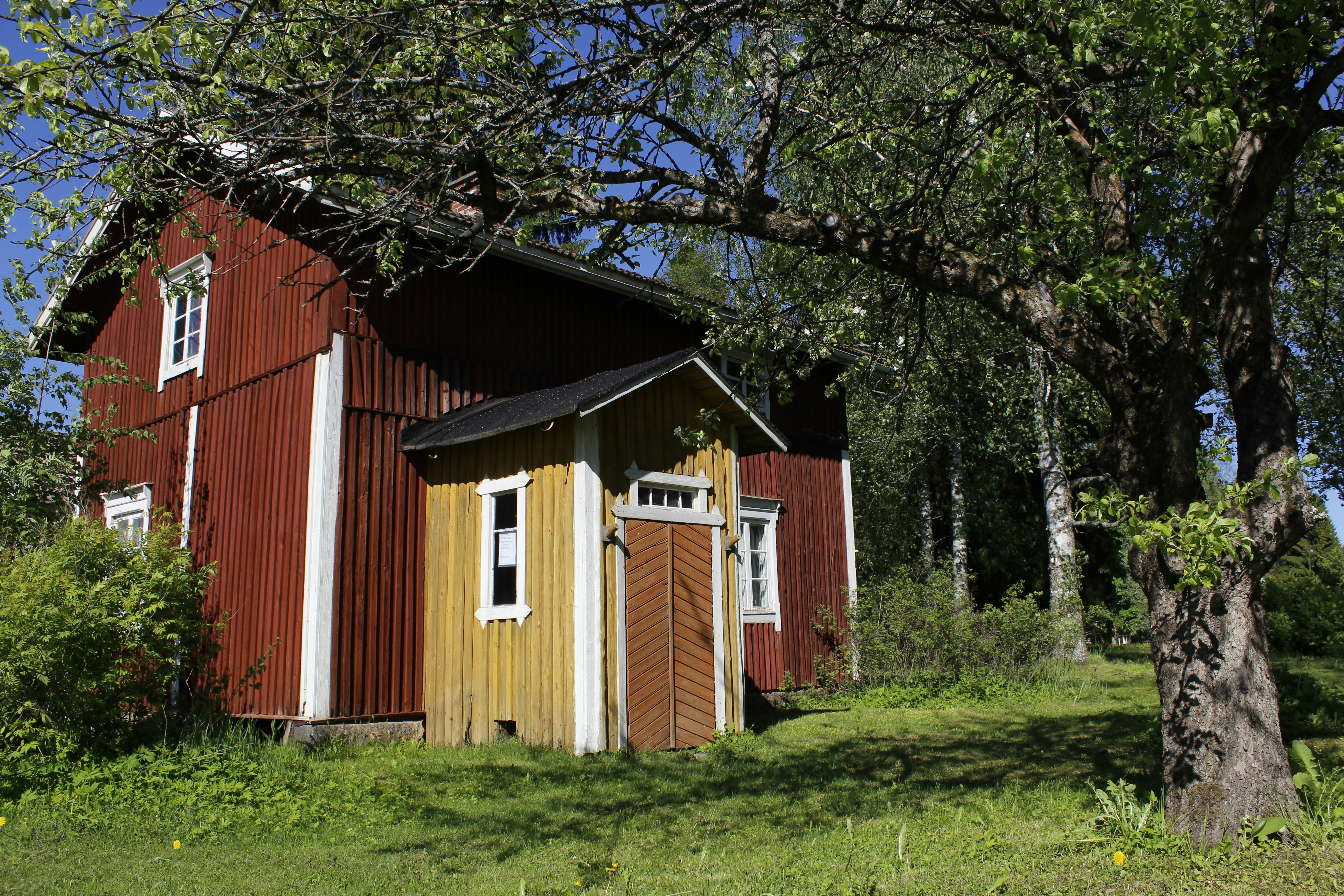 A quaint wooden house with red and yellow accents nestled in a lush green landscape, framed by a large tree. The scene evokes a sense of tranquility and rustic charm.