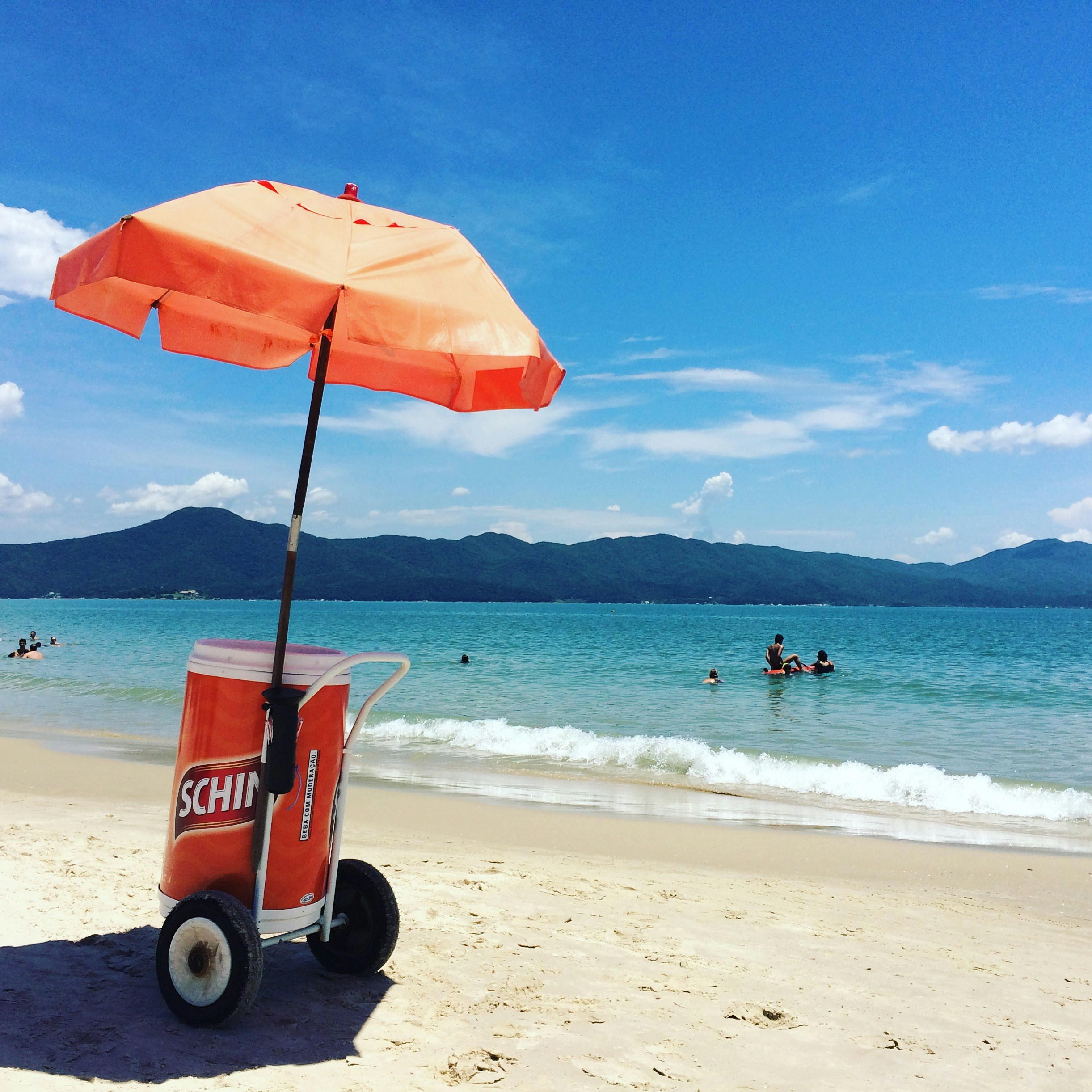 Bright orange beach umbrella stands beside a rolling cart on sandy shore, with gentle waves lapping at the beach and people enjoying the water in the background.