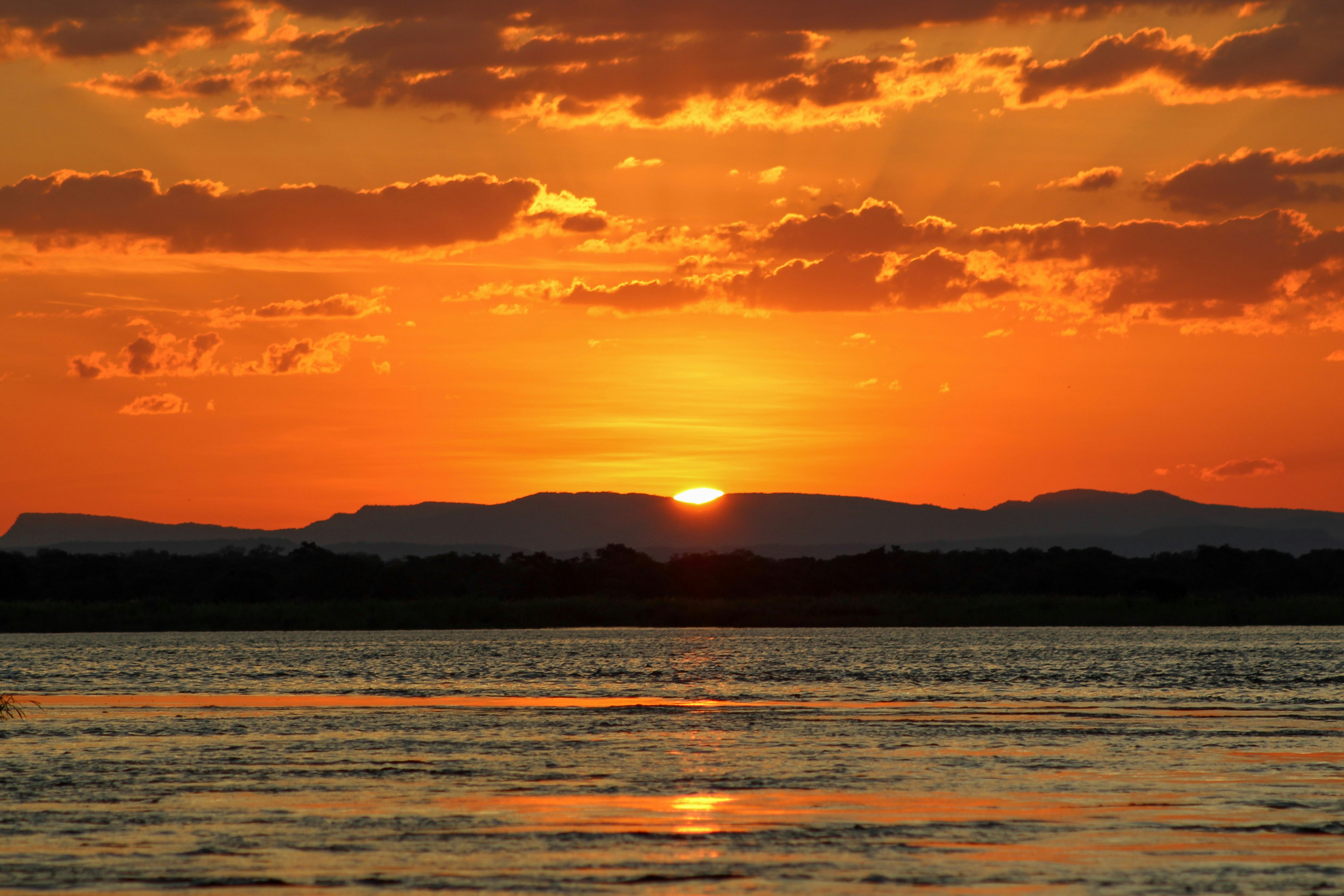 Vibrant sunset casting warm hues over tranquil waters, with silhouetted mountains in the background. The scene evokes a sense of peace and reflection.