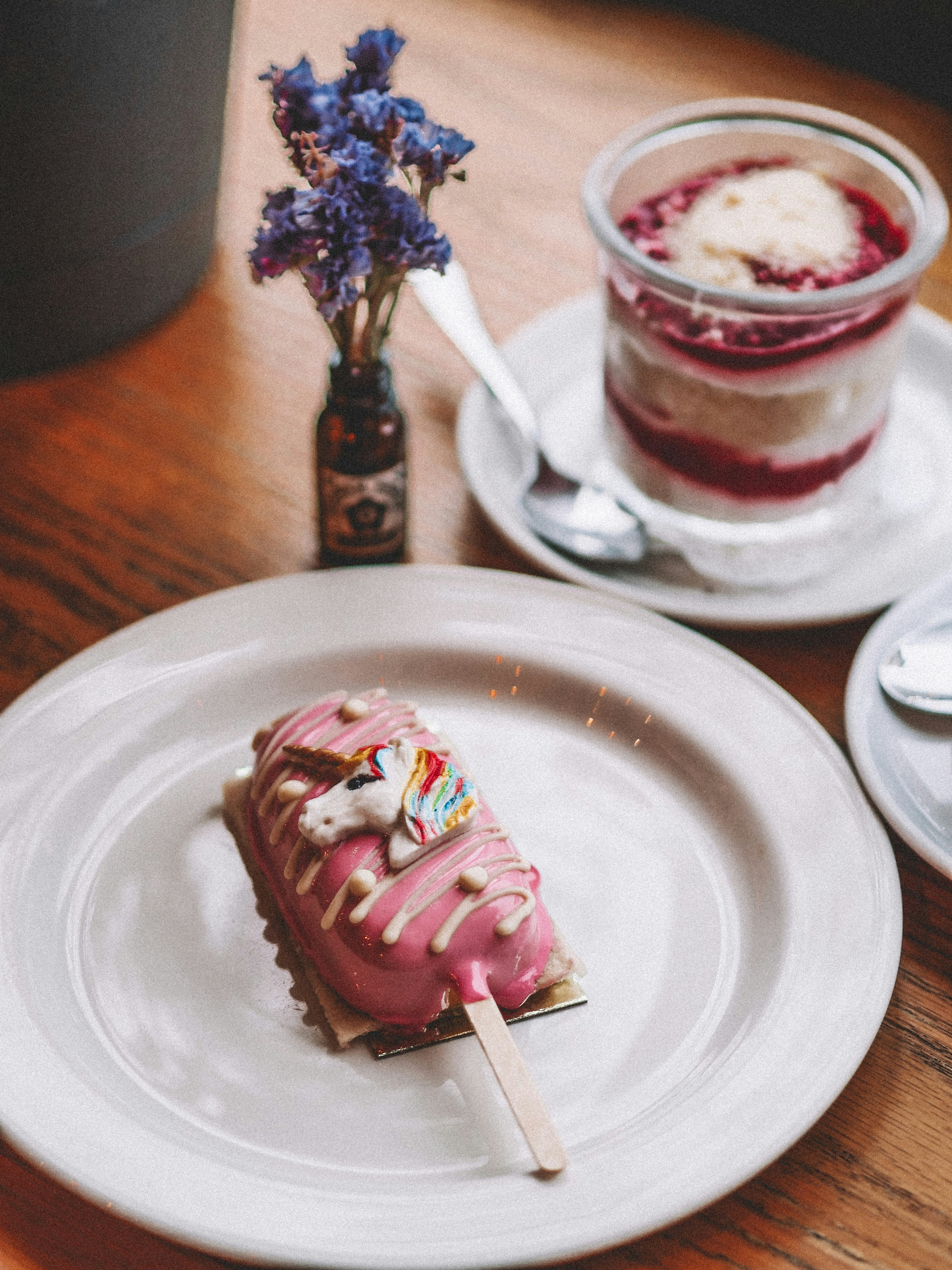 Colorful unicorn-themed ice cream bar on a plate, accompanied by a layered dessert and a sprig of flowers. Perfect for a delightful afternoon indulgence.