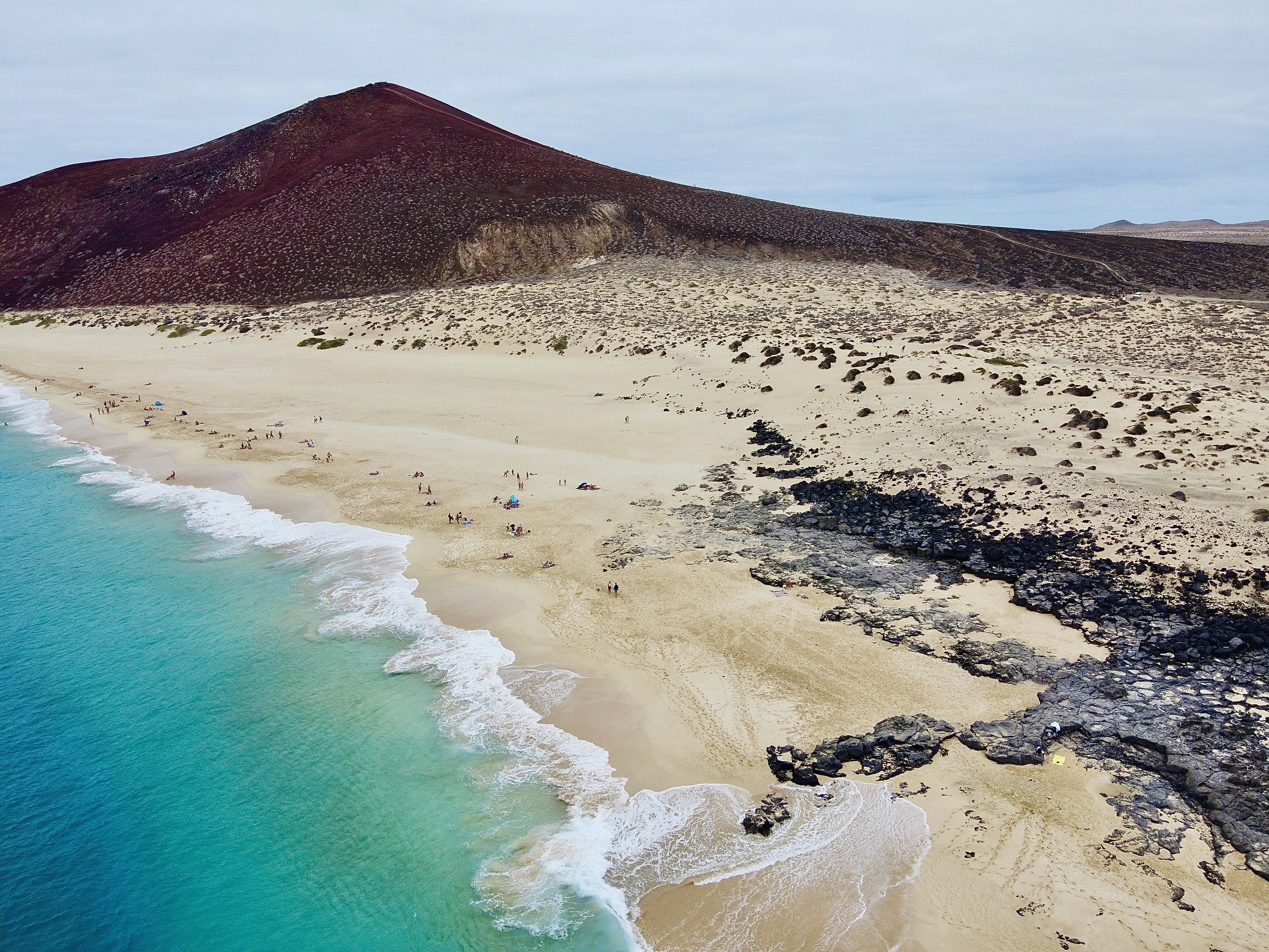 Aerial view of a serene beach with golden sands and turquoise waters, framed by a volcanic hill and rocky coastline. People can be seen enjoying the beach.