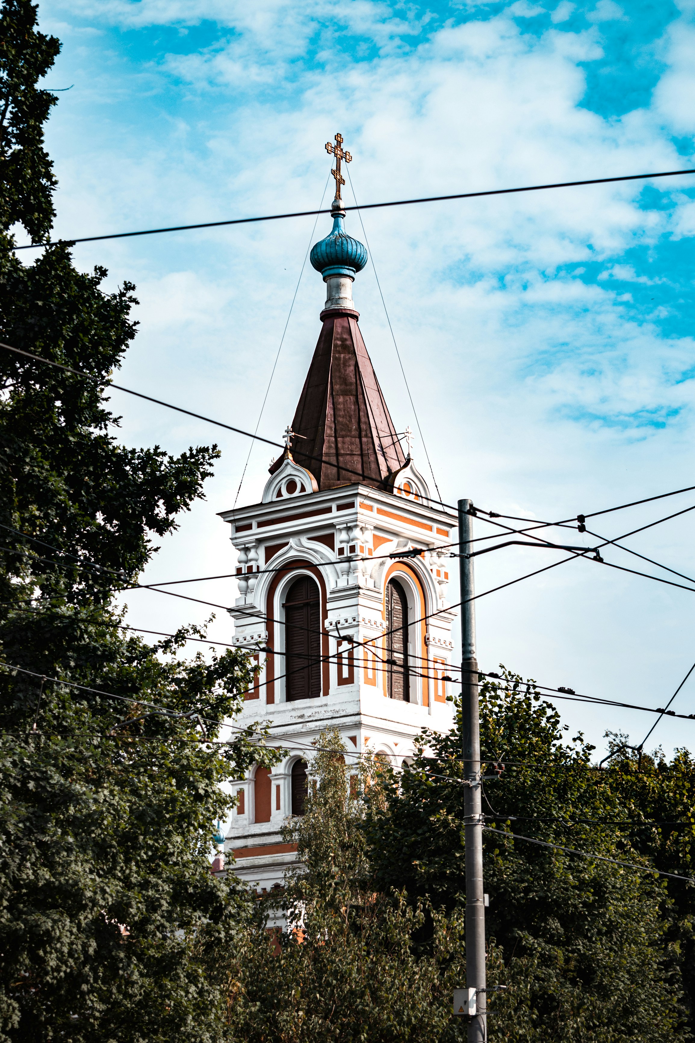 A charming church steeple rises above lush greenery, framed by power lines against a bright sky.