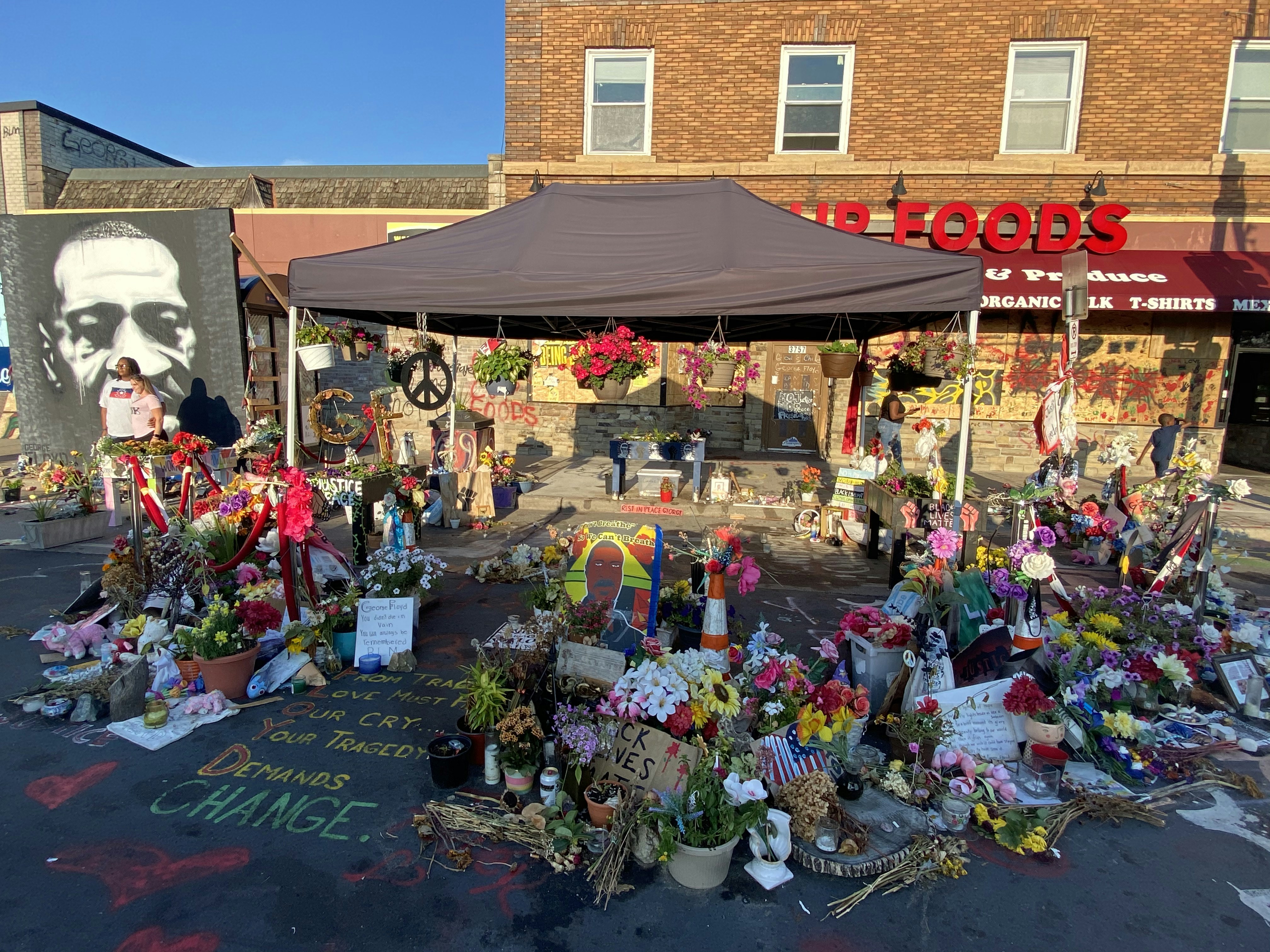 Memorial site adorned with flowers and tributes under a tent in front of a mural.