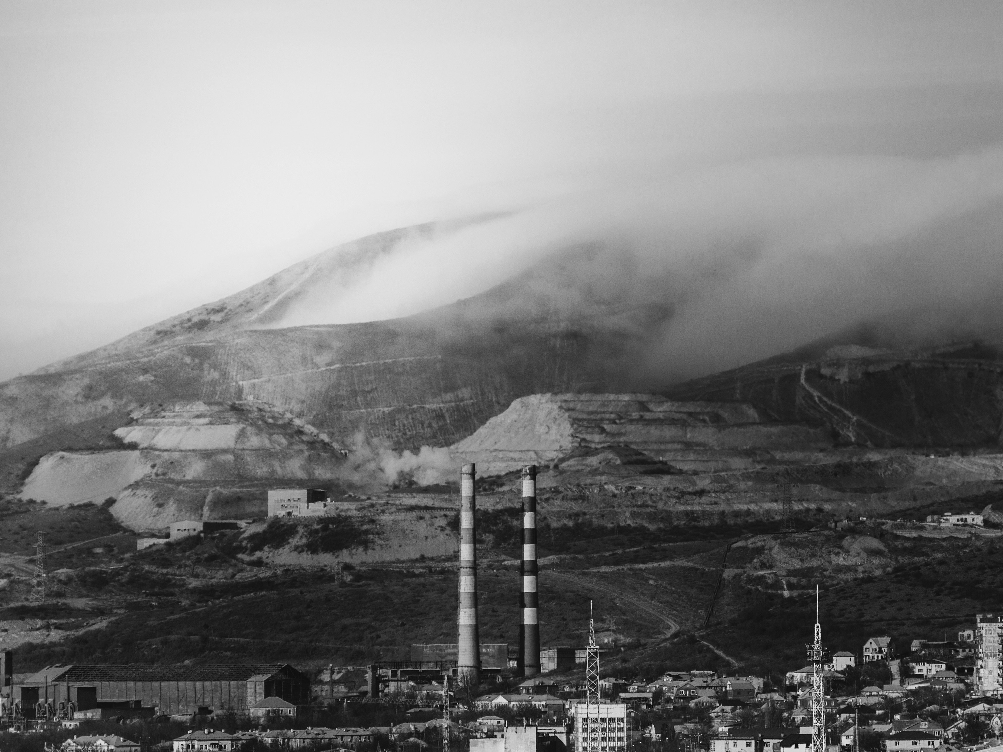 Industrial smokestacks rise against a misty mountain backdrop, shrouded in clouds and revealing the stark contrast between nature and human activity.