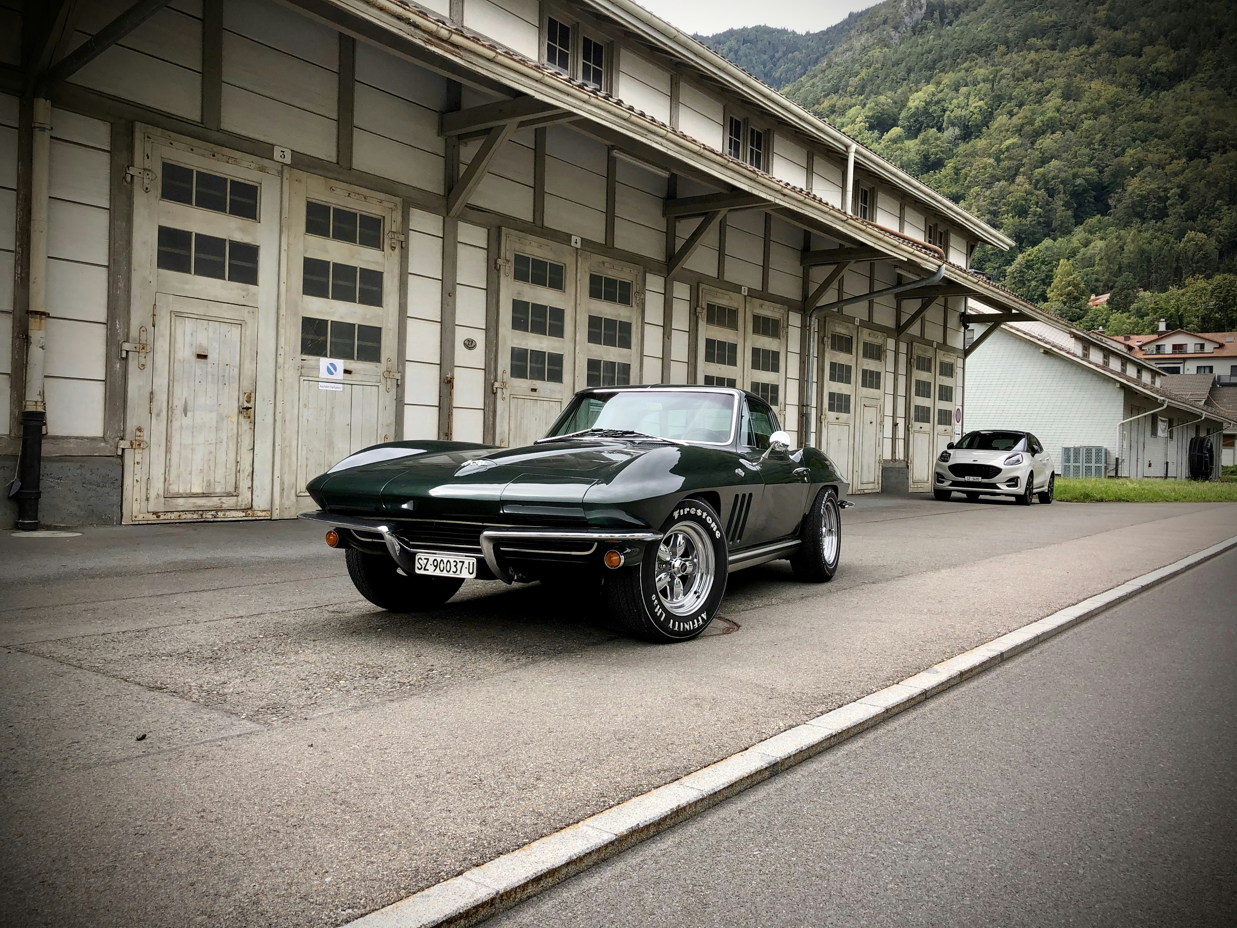 Vintage Chevrolet Corvette StingRay parked in front of an industrial building with rolling hills in the background.