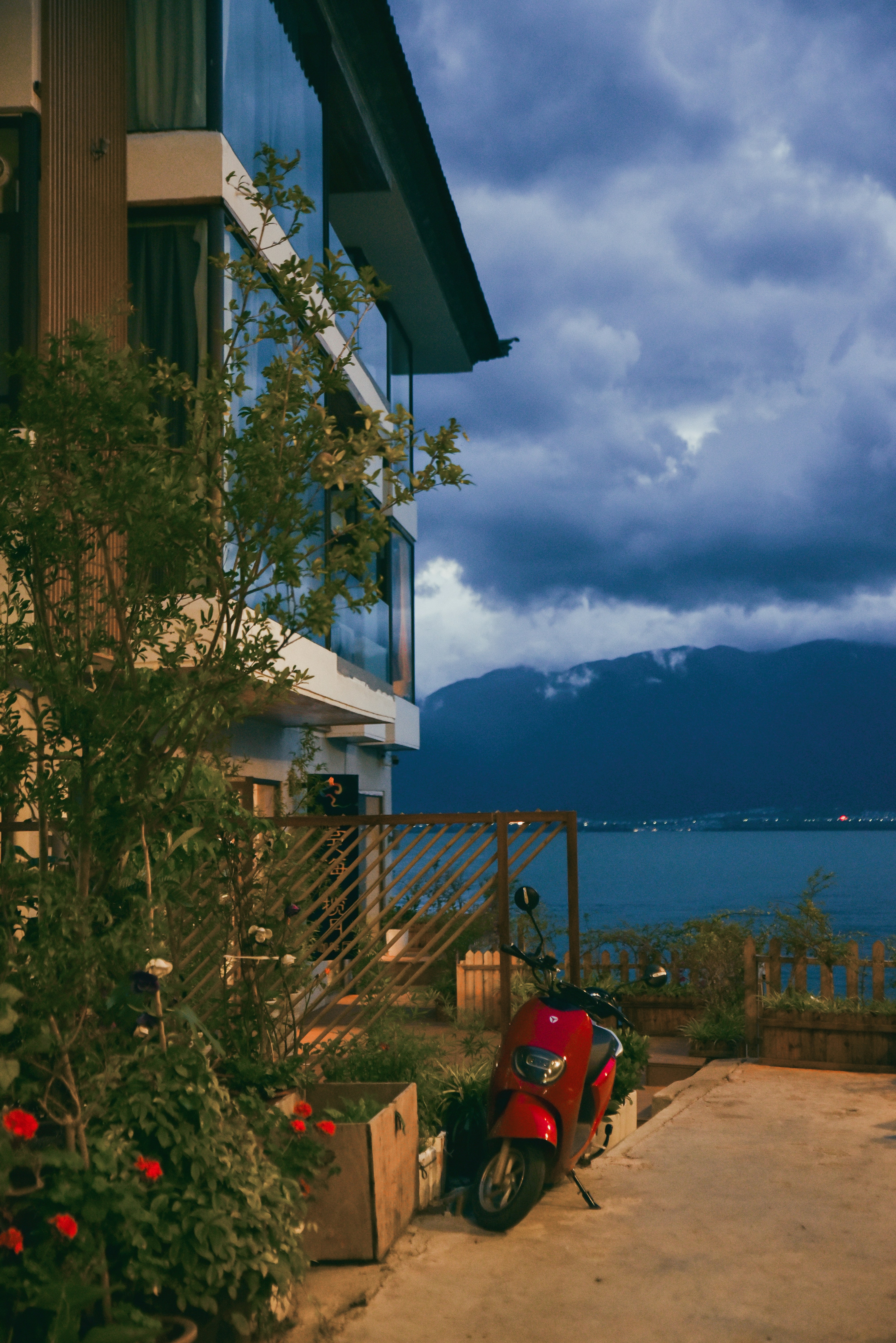 A vibrant red scooter parked beside a lakeside garden, framed by lush greenery and a backdrop of dramatic clouds over distant mountains.