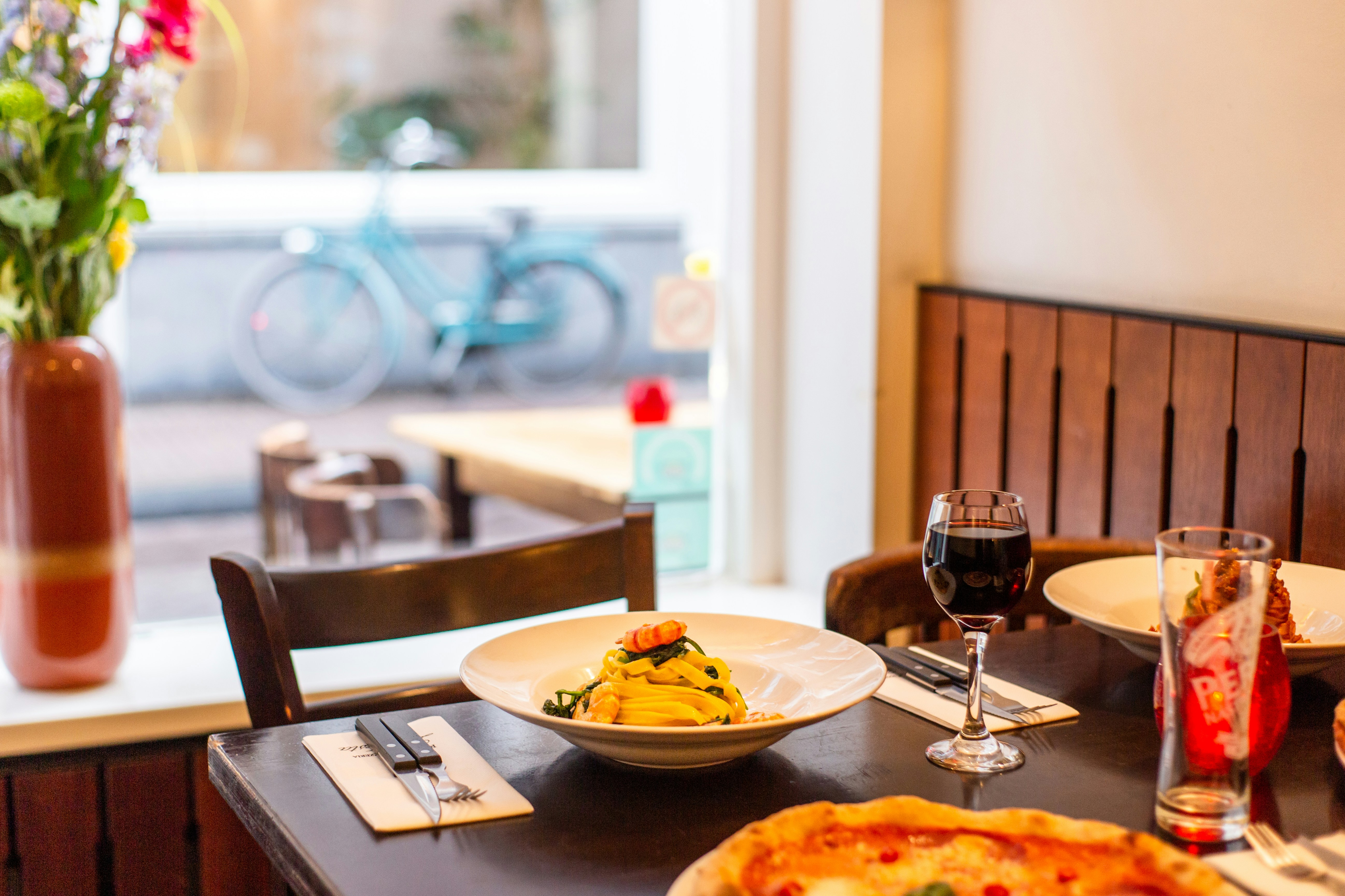 Pasta and pizza on a wooden table in a cozy café setting, with a bicycle visible through the window.