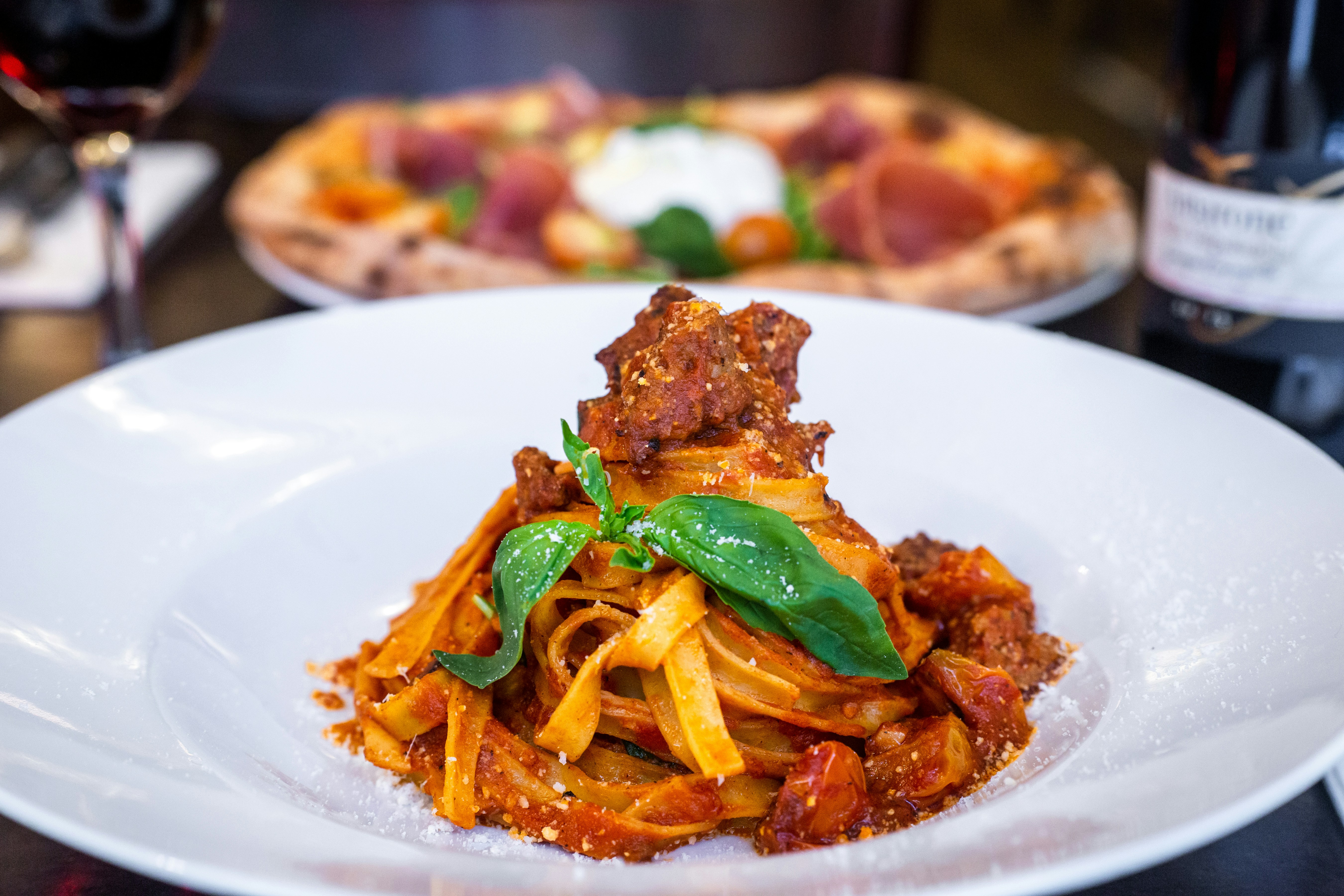 Plate of tagliatelle pasta with basil garnish in focus, with a pizza and wine in the background.