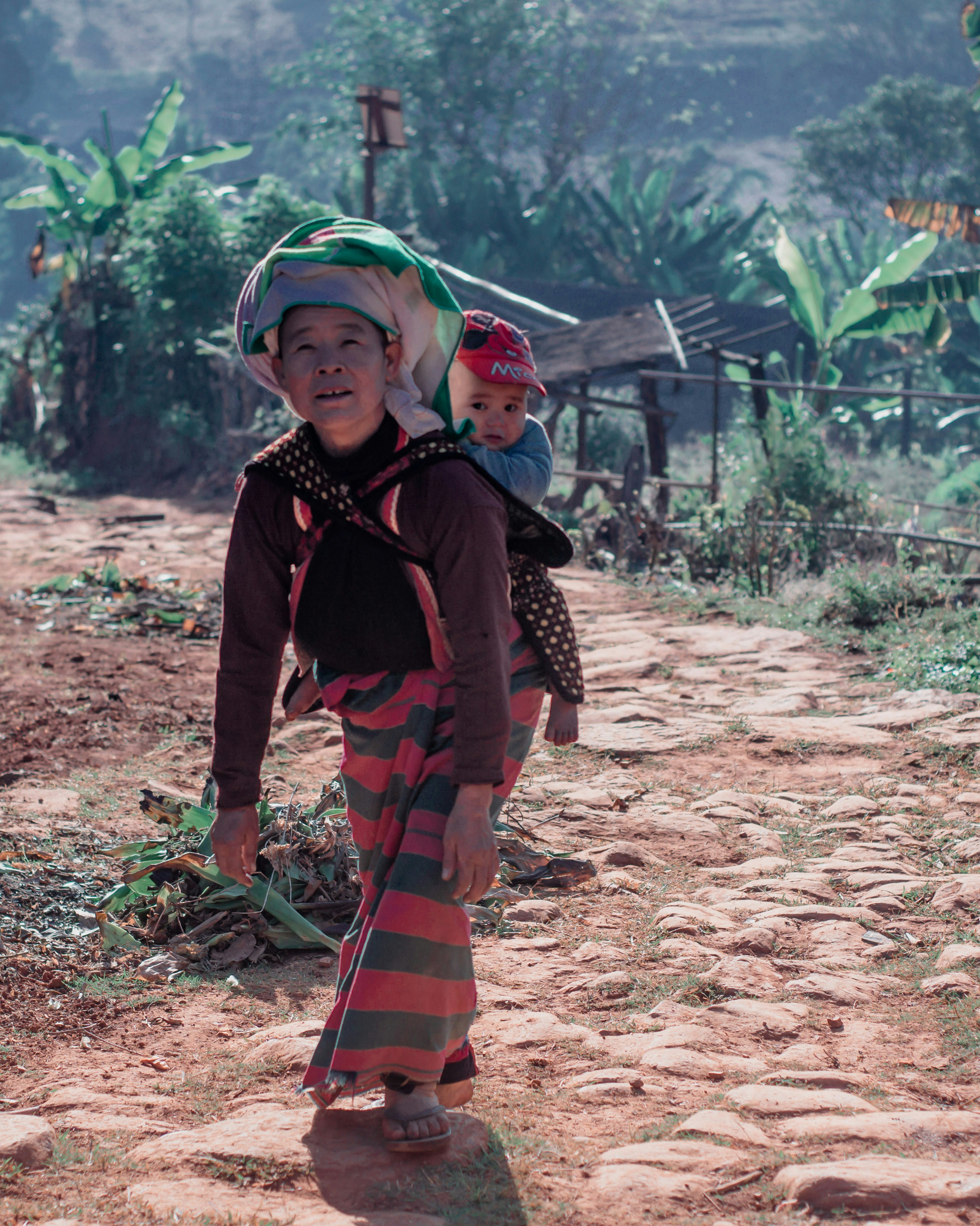Woman carrying a child on her back while walking along a dirt path in a lush, green landscape.