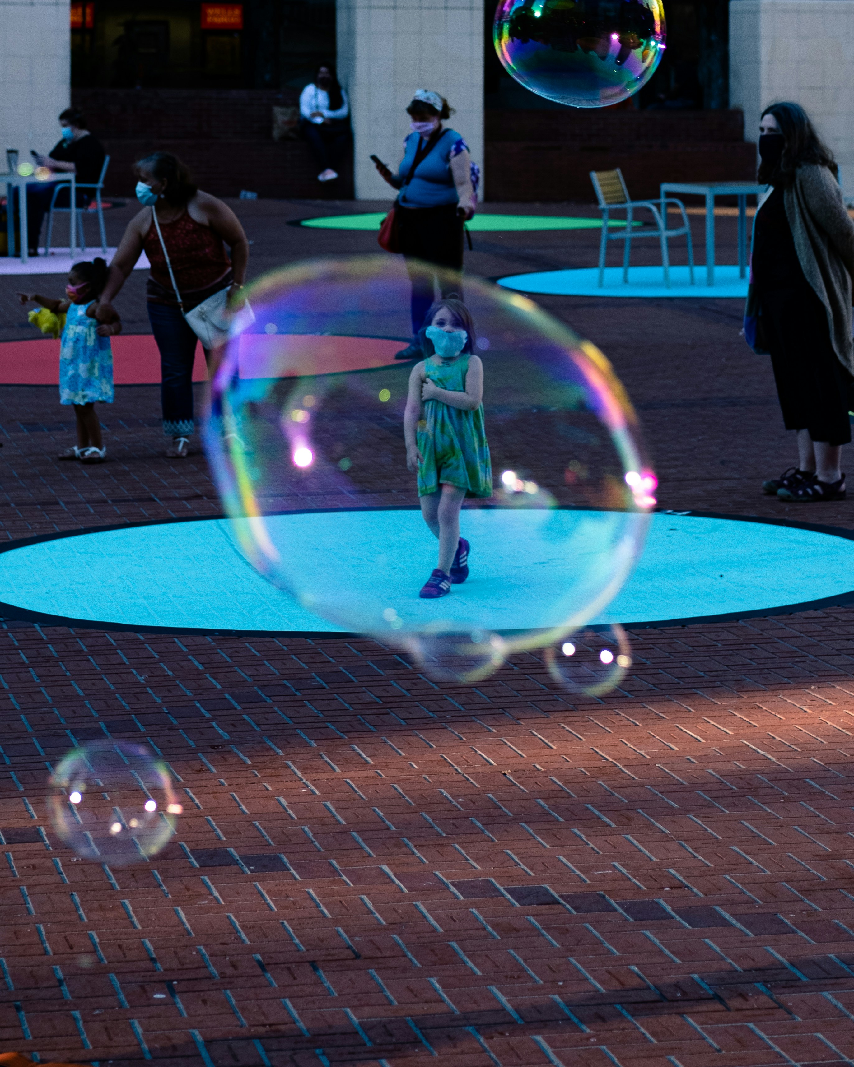 A young girl in a green dress stands amidst large soap bubbles, surrounded by people in a vibrant outdoor setting. The scene captures a playful and joyful atmosphere.