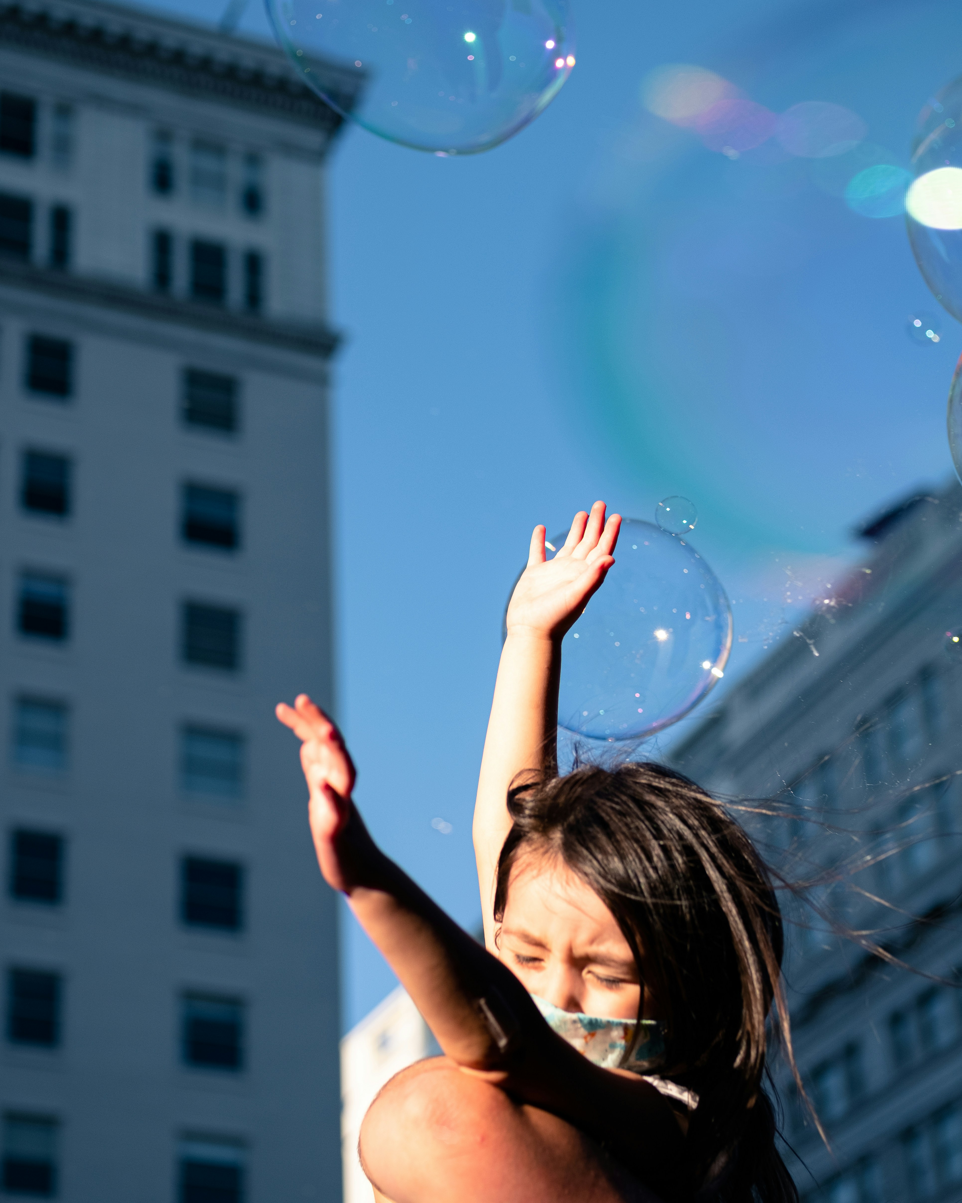 Young child with a mask reaching for floating bubbles against a backdrop of urban architecture.
