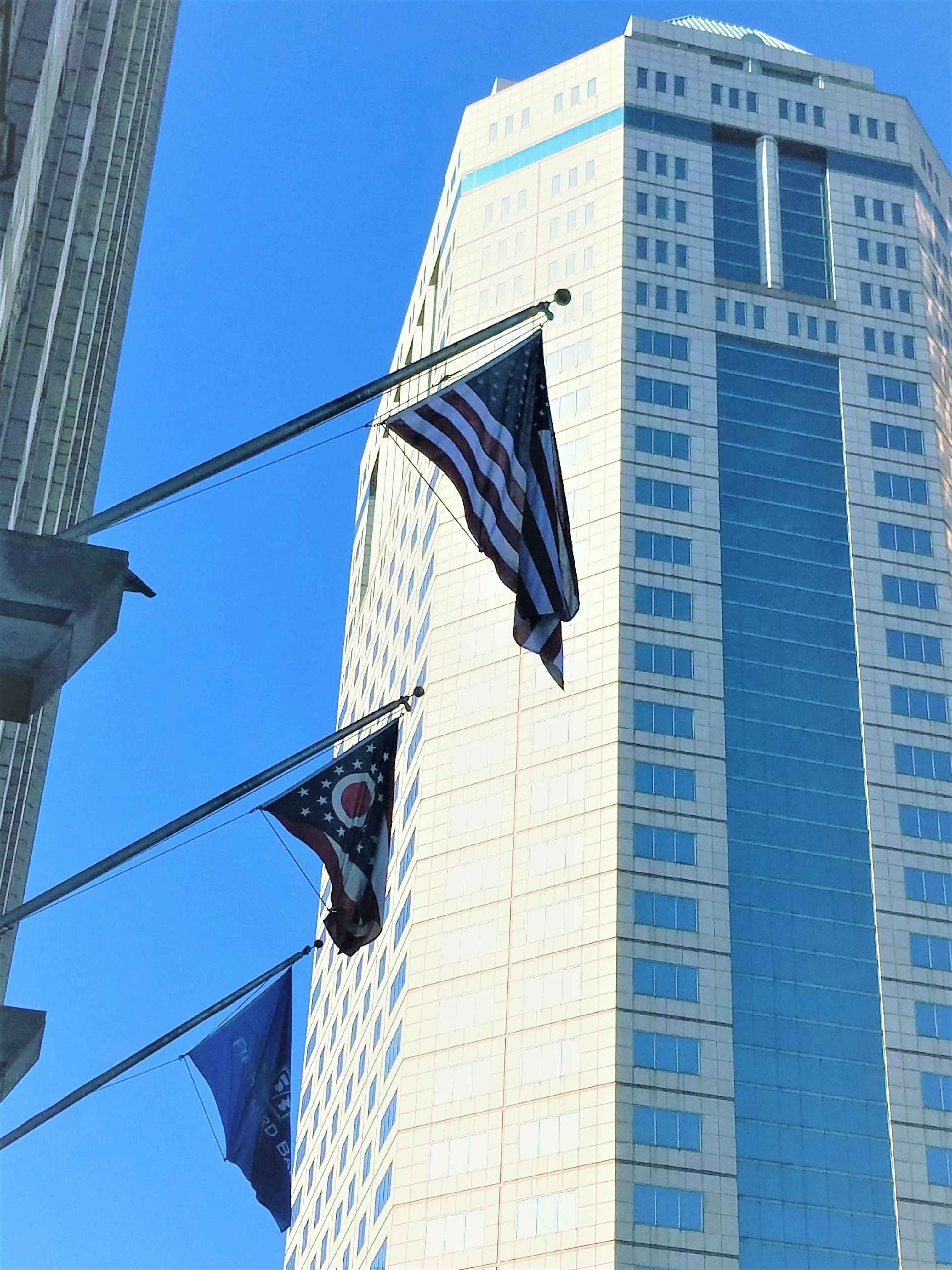 Three flags fluttering in the breeze against a backdrop of a modern skyscraper and clear blue sky.