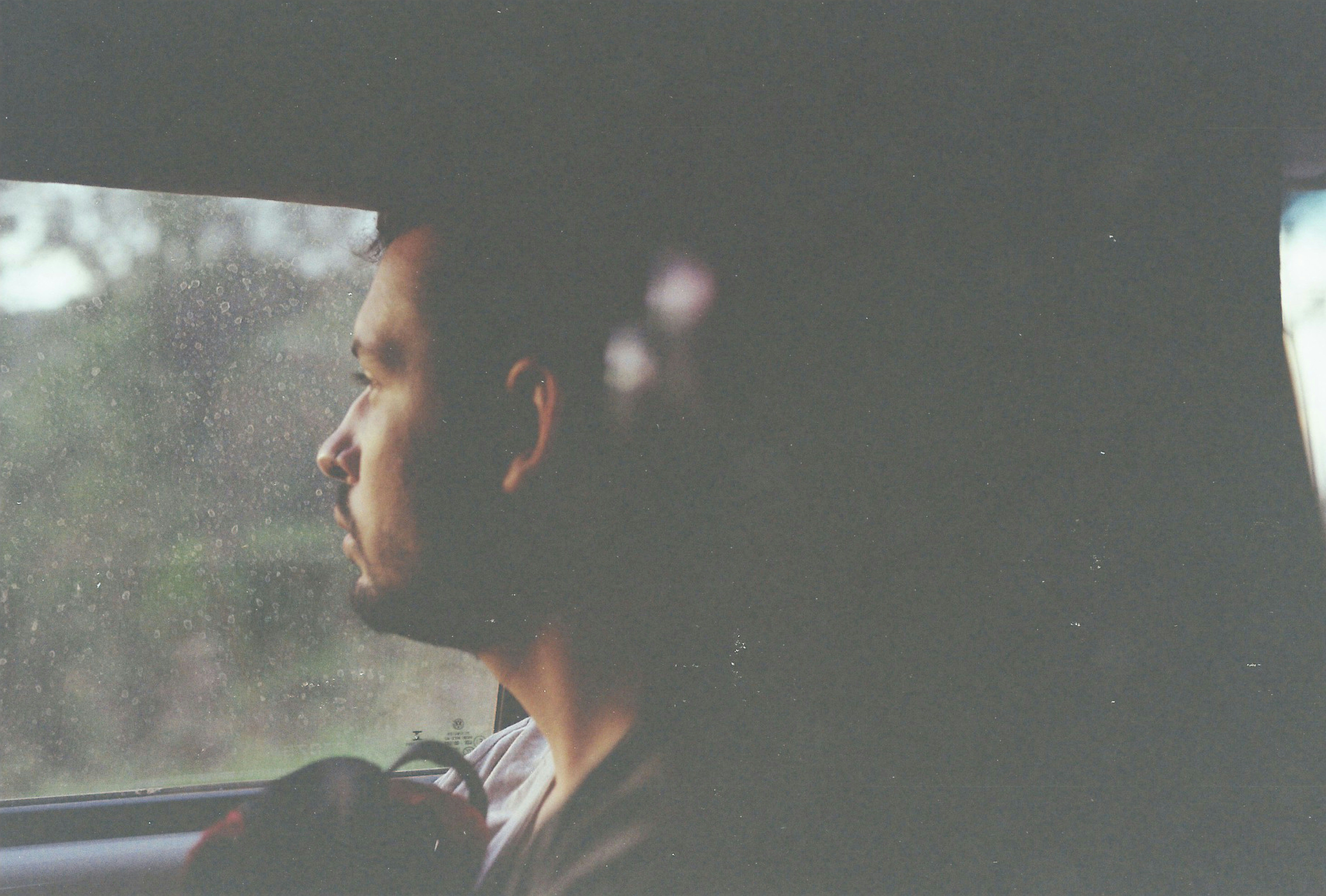 A solitary man in profile sits in a dim car, gazing left with rain-speckled glass and a softly blurred exterior.