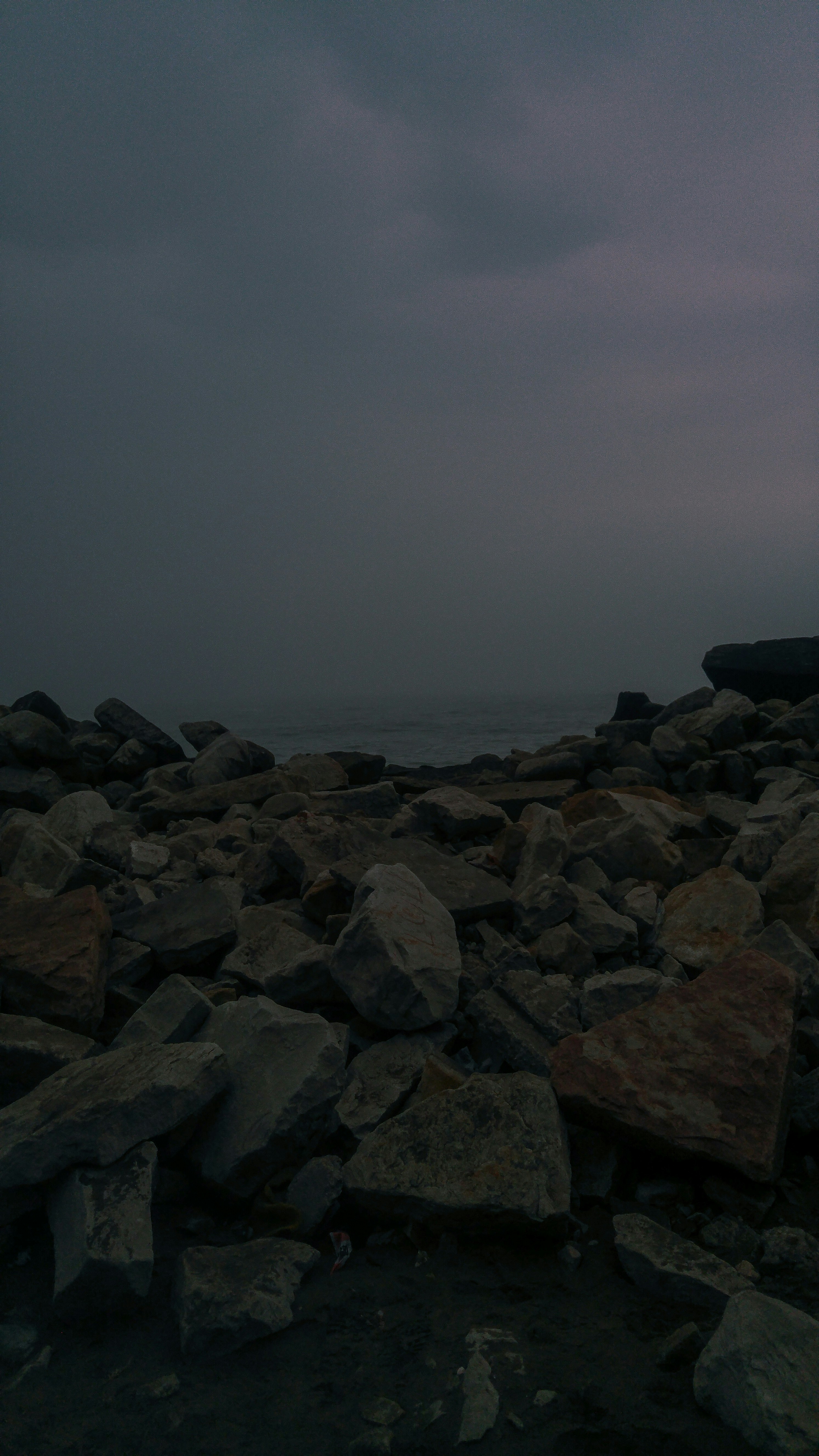 Jagged rocks scatter along the shoreline under a moody sky, hinting at the tumultuous sea beyond. The atmosphere evokes a sense of solitude and contemplation.