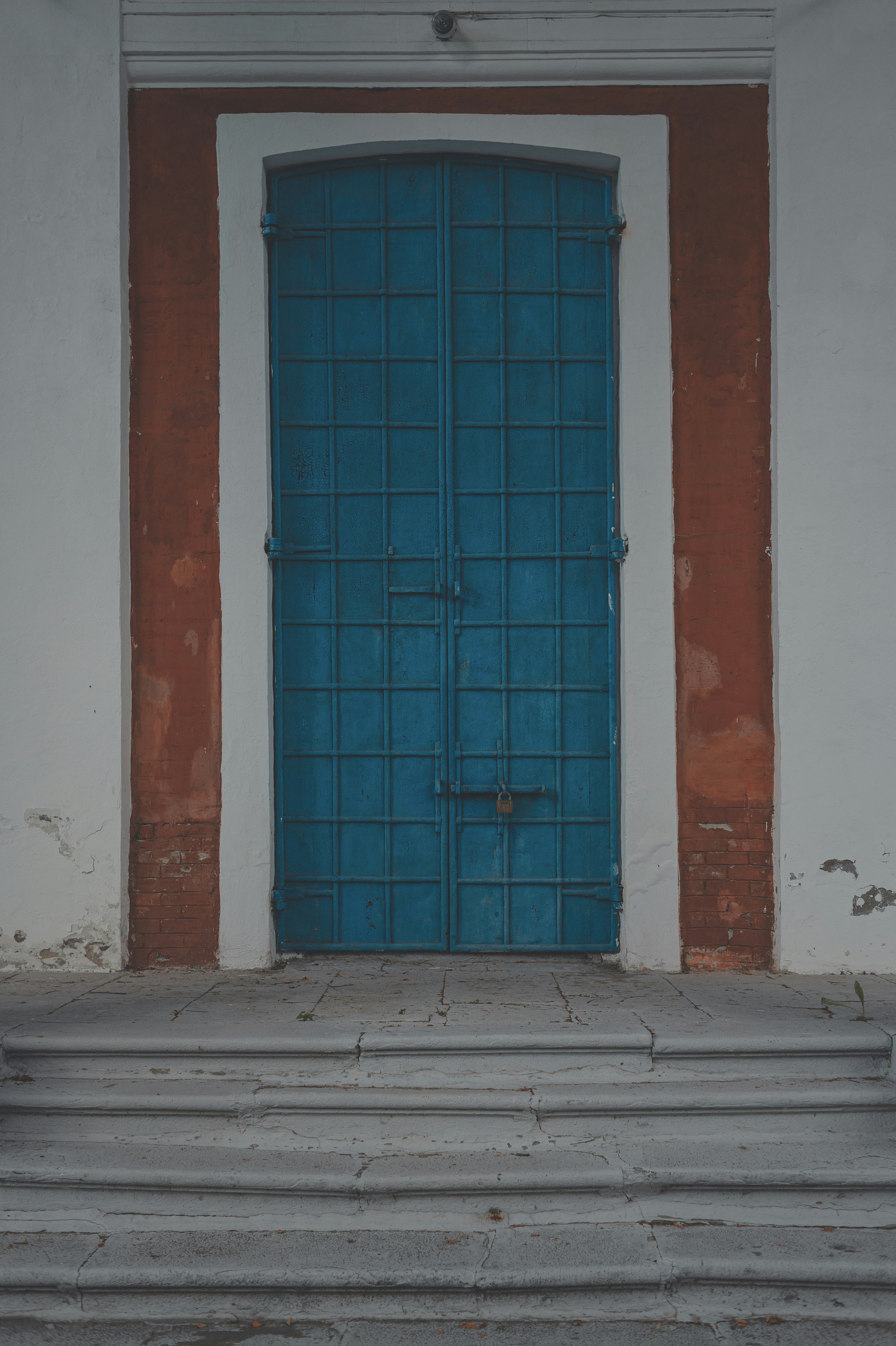 A vibrant blue door with a lock stands against a weathered white wall, framed by rustic red accents and stone steps.