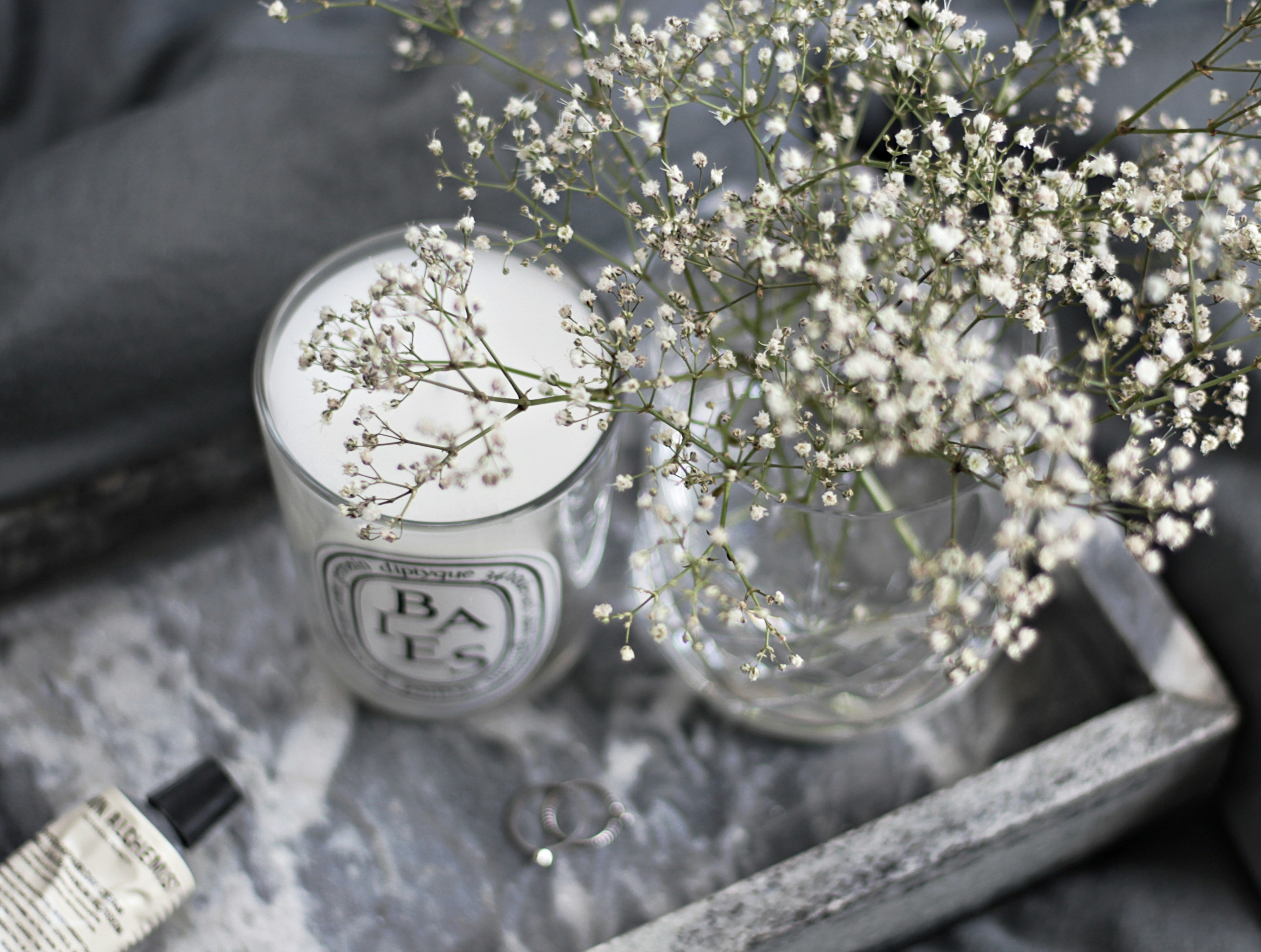 Delicate white flowers arranged in a glass vase beside a scented candle and a small tube of cream on a textured tray.
