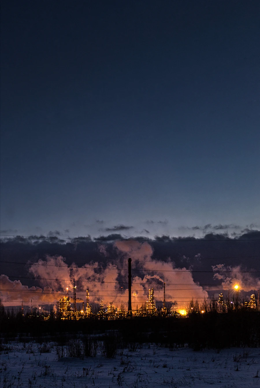 Illuminated oil refinery silhouetted against a twilight sky, with steam rising from chimneys and a snowy foreground.