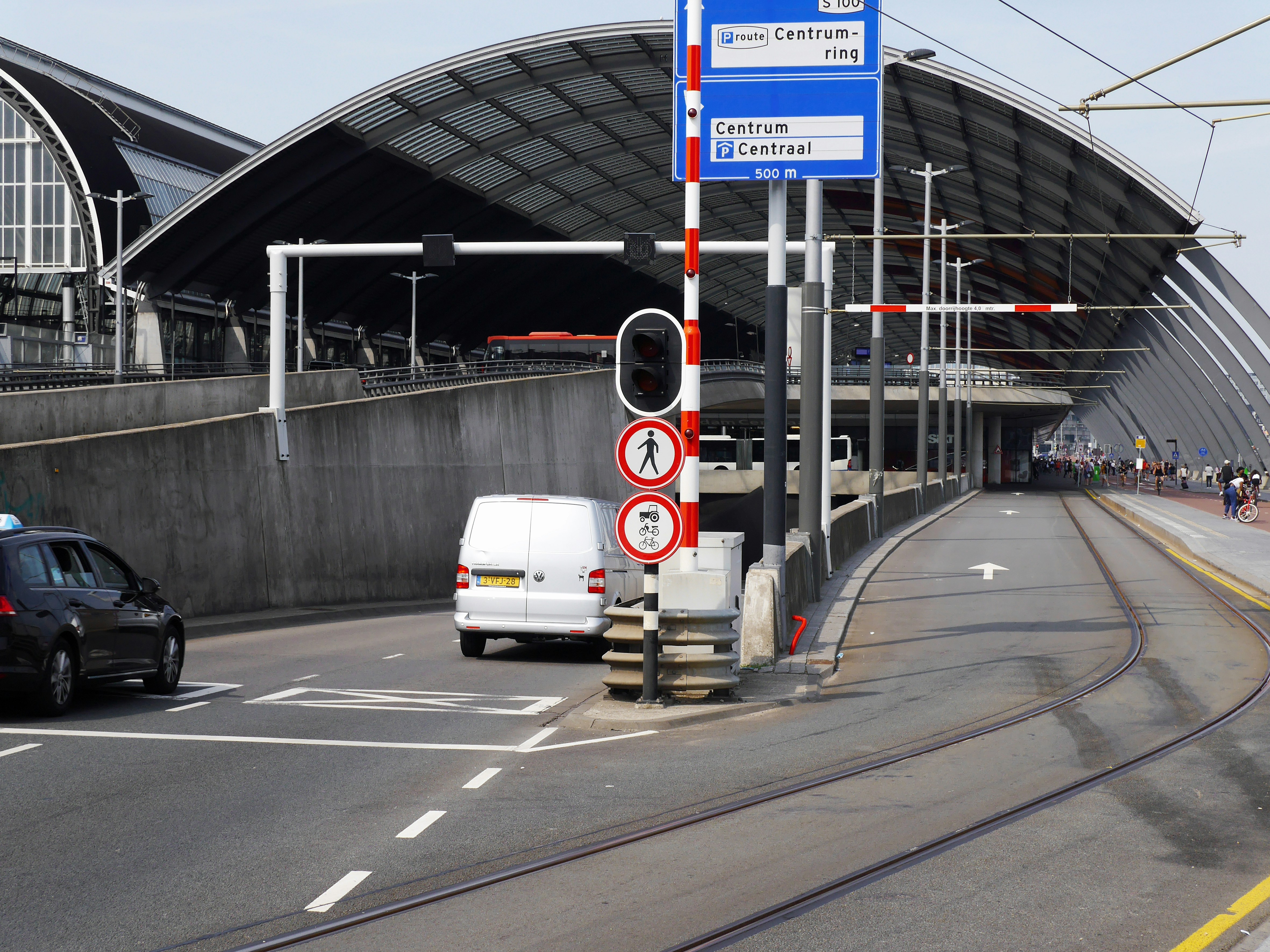 White van on a quiet urban street in front of a modern curved-roof train station. Tram tracks curve along the right while overhead signs and poles frame the scene.