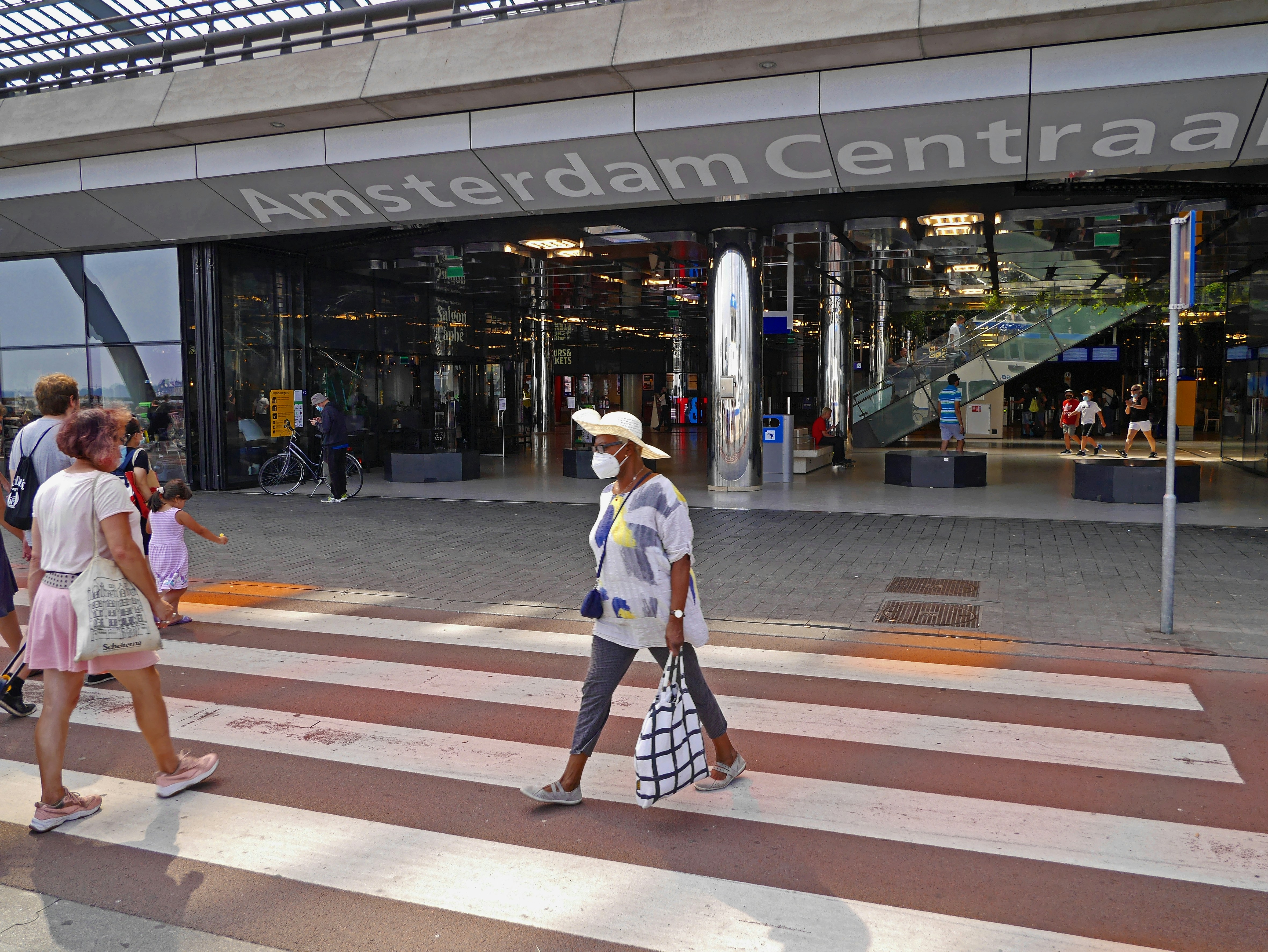 Pedestrians cross the iconic striped crosswalk in front of Amsterdam Centraal station, showcasing the vibrant city life and modern architecture.