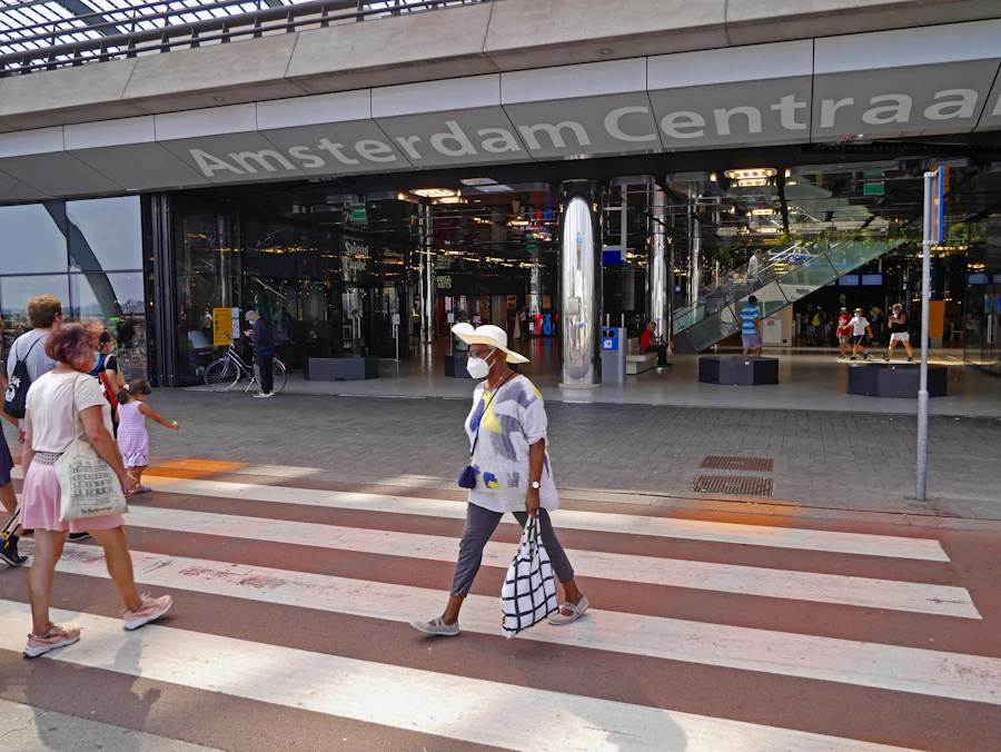 People crossing a wide striped crosswalk in front of the Amsterdam Centraal station entrance, with glass reflections and busy foot traffic. A masked woman in a hat walks through the center foreground.