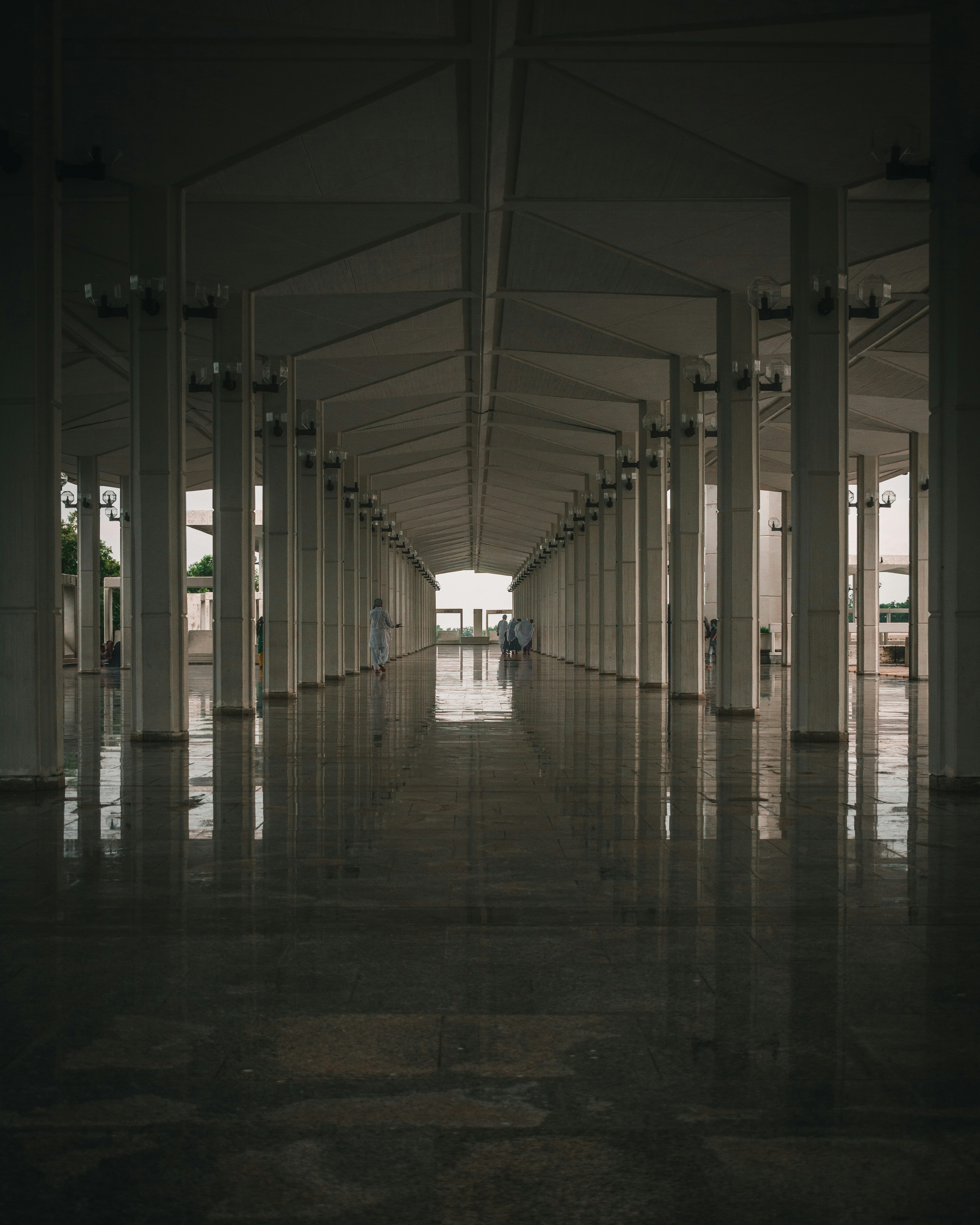 A spacious corridor lined with tall columns and reflective flooring, leading to an open entrance. The scene captures the tranquility of architectural design.