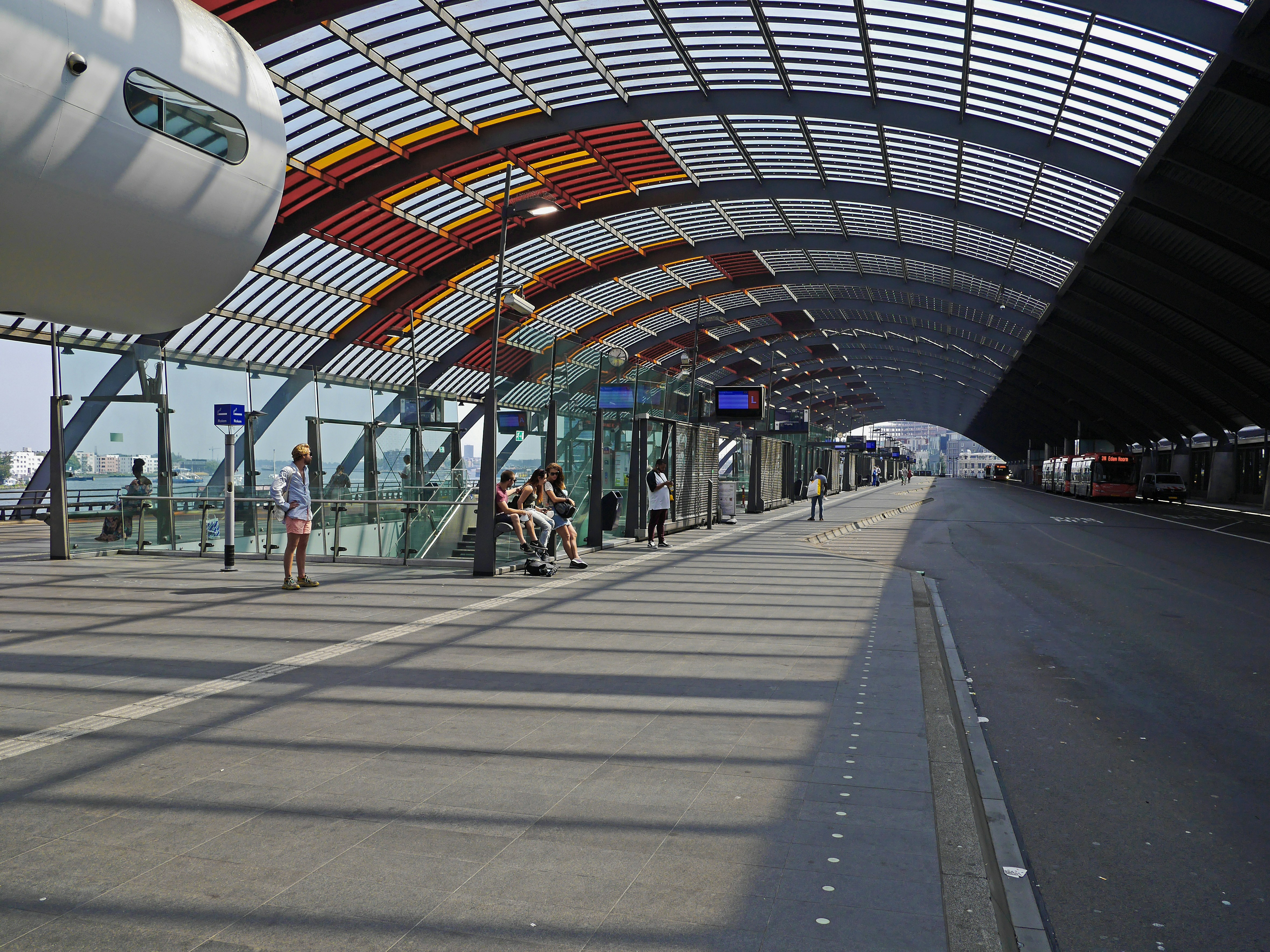 Sleek transit station interior showcasing a striking roof design and spacious waiting area, with passengers engaging in various activities.