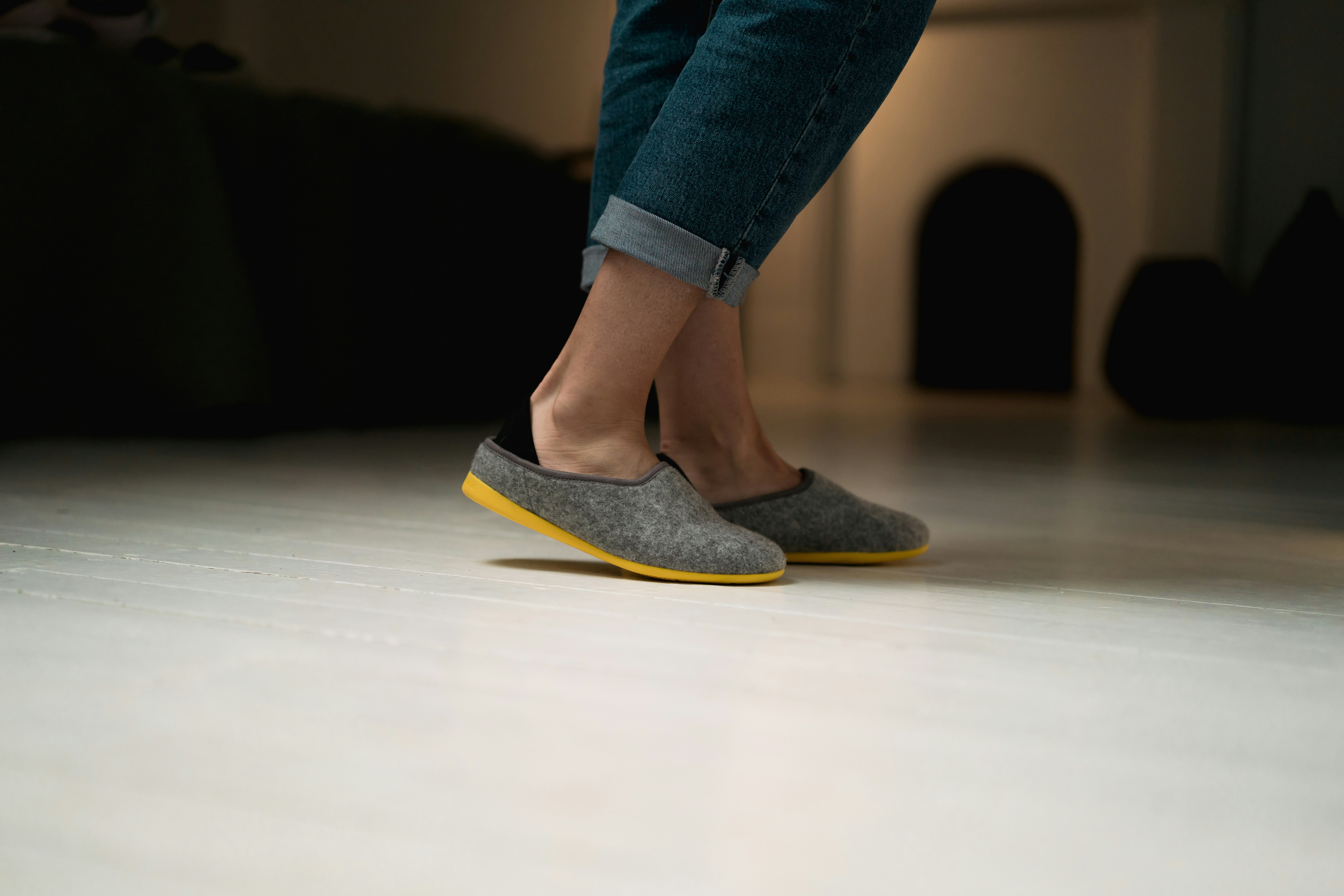 Gray slippers with yellow soles, worn by a person, resting on a light wooden floor in a softly lit interior space.
