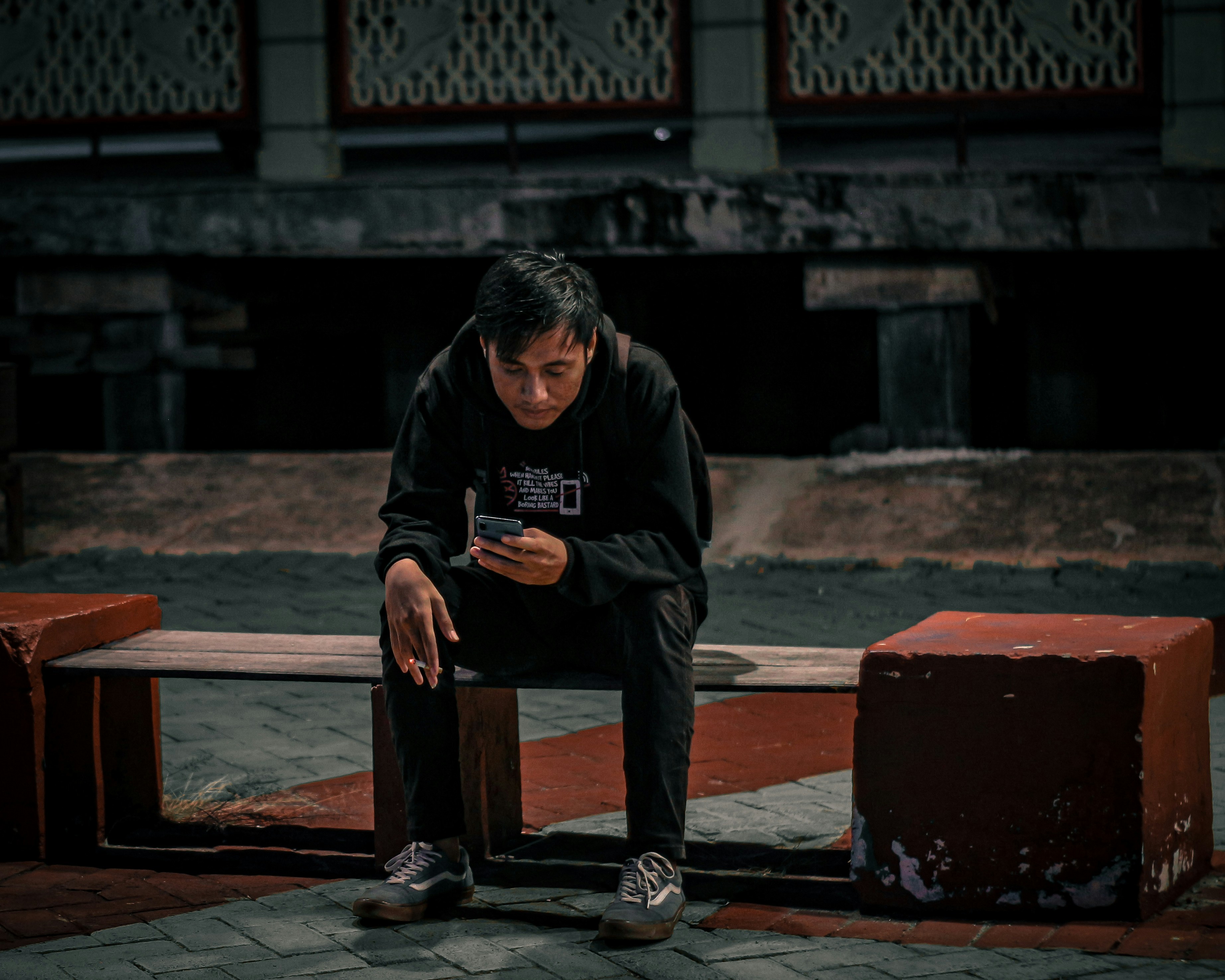 A young man sitting on a bench, absorbed in his smartphone, surrounded by urban architecture at night.