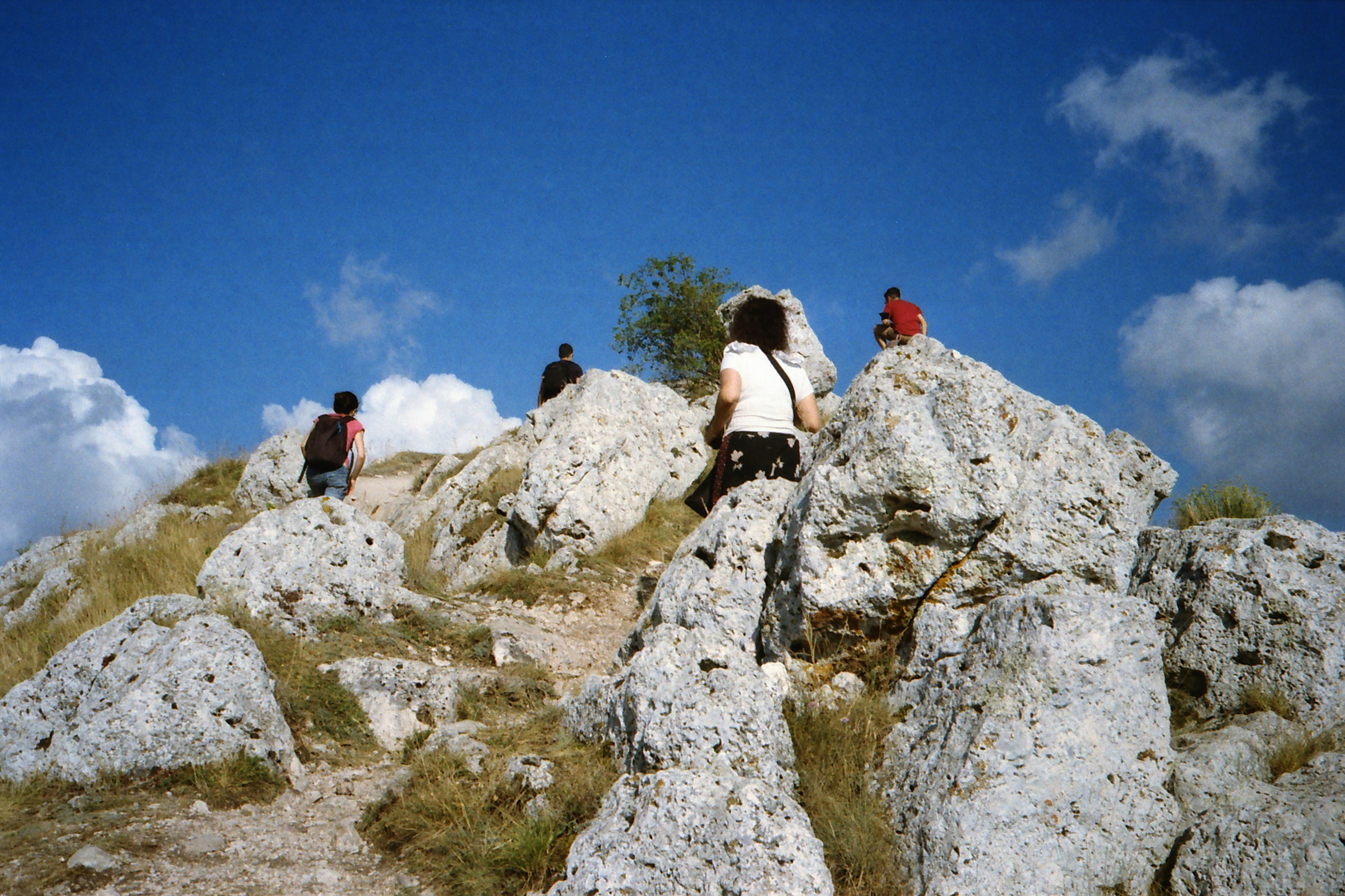 Hikers navigating rocky paths on a hillside under a blue sky with fluffy clouds.